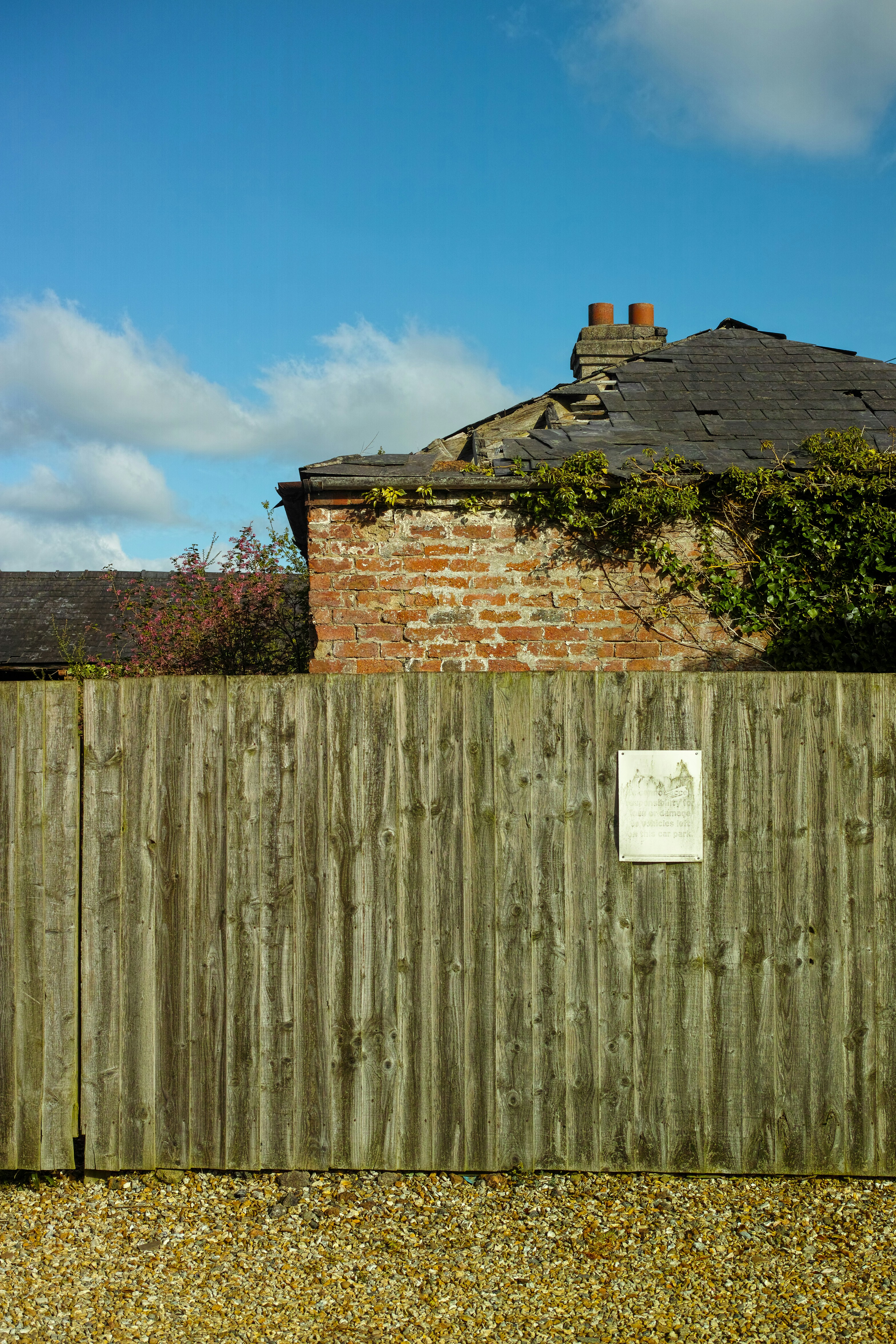 A weathered wooden fence dominates the foreground while a brick house peeks over the top under a bright blue sky. A small white notice is affixed to the fence, drawing the eye to a subtle focal point.