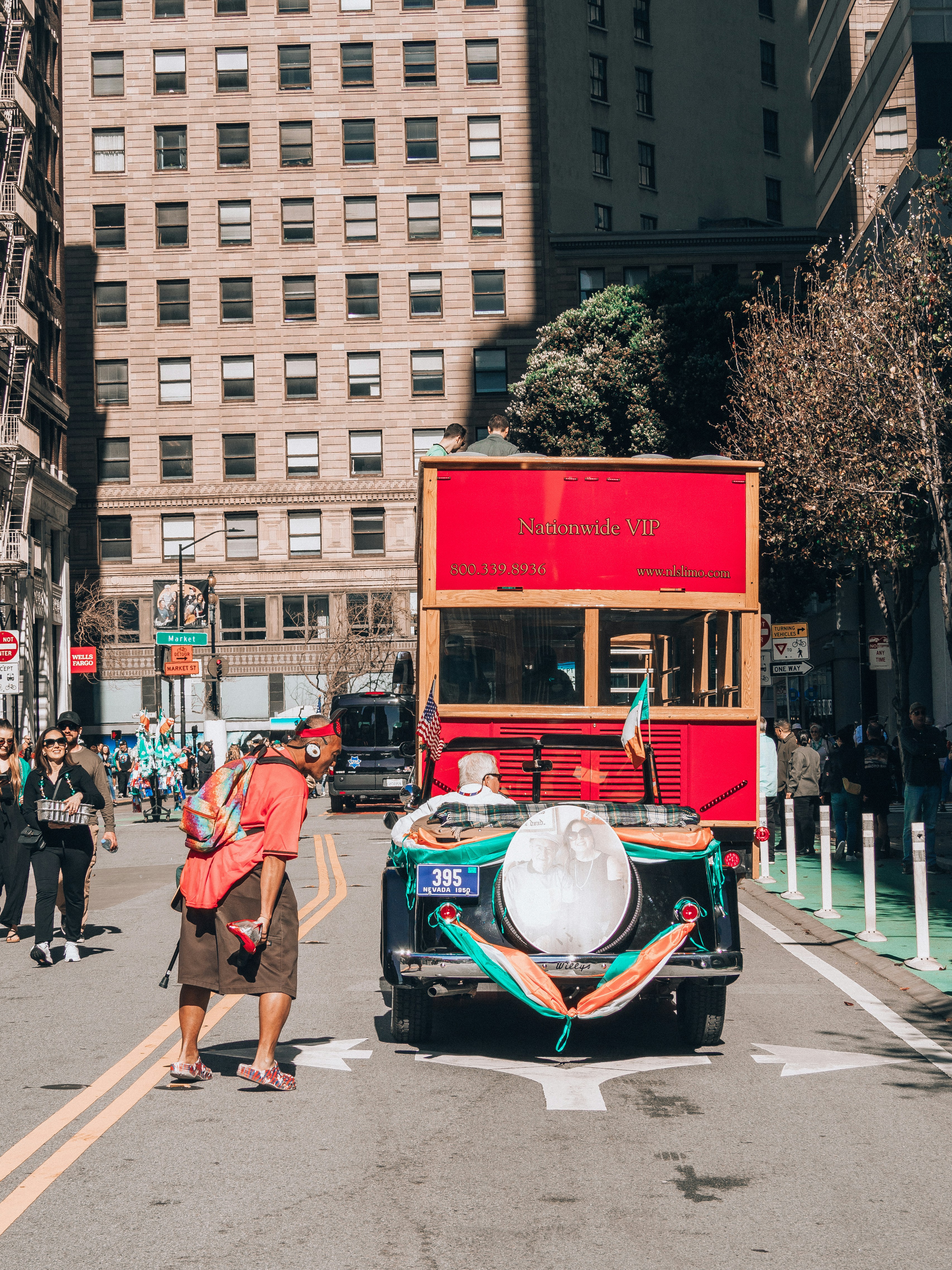 A vintage car leads a parade through a city street with a red double-decker bus in the background and pedestrians on either side. A skyline of tall office buildings looms as the scene captures motion and color in a street celebration.