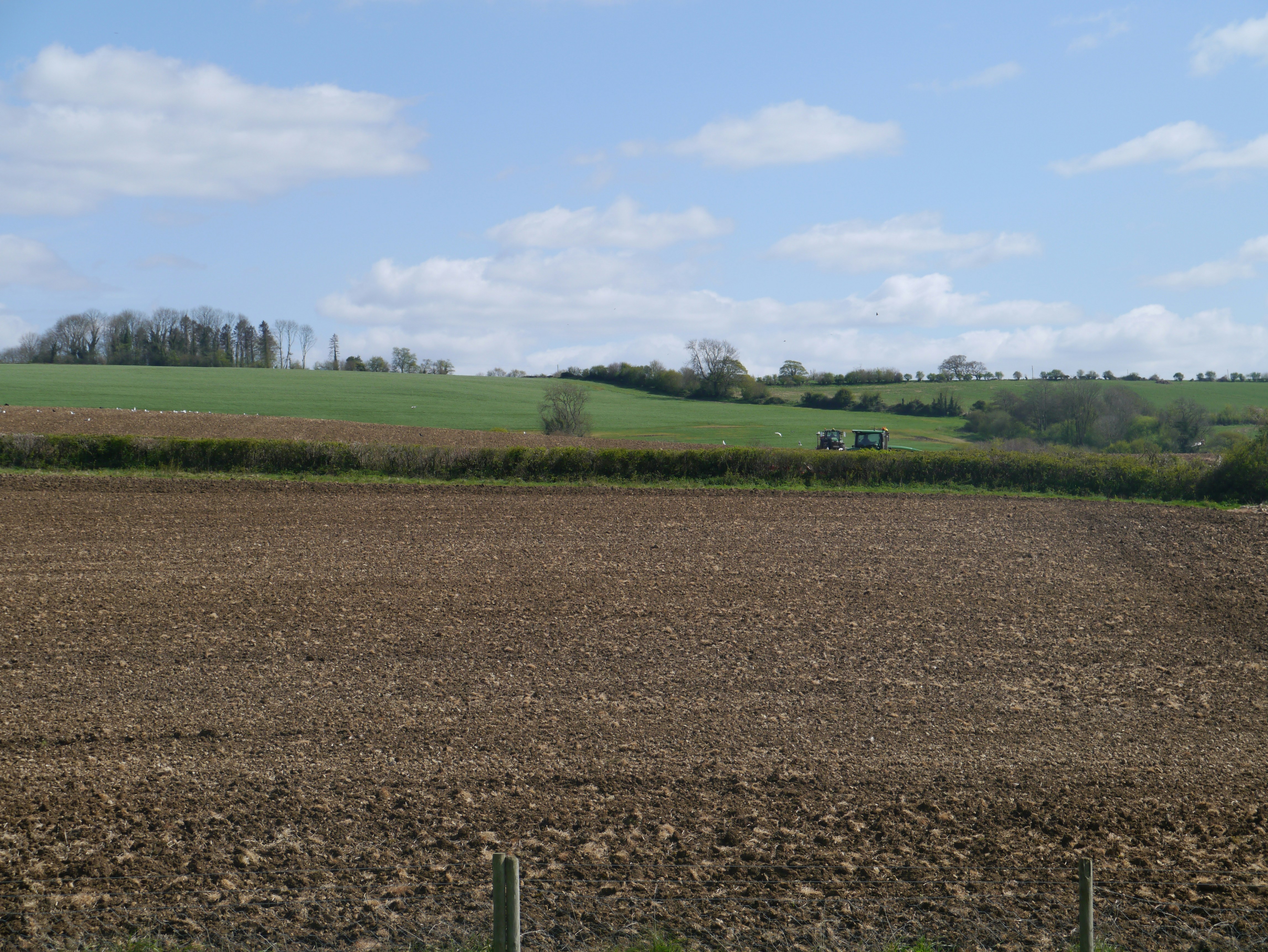 Landscape photograph of a rural field under a bright blue sky, with a small tractor on the distant horizon.