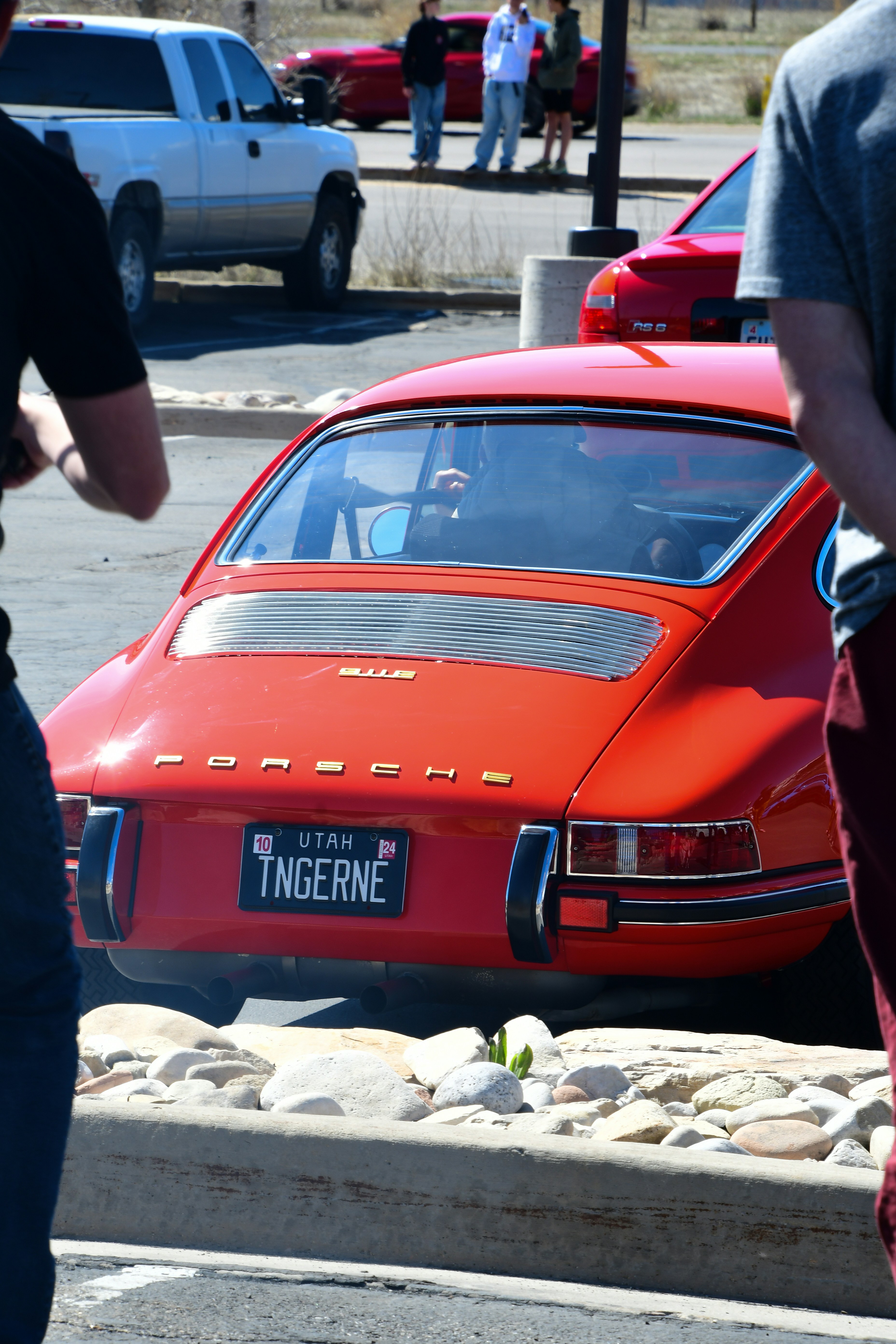 Scarlet Porsche 911 at a sunlit parking lot gathering, framed by onlookers