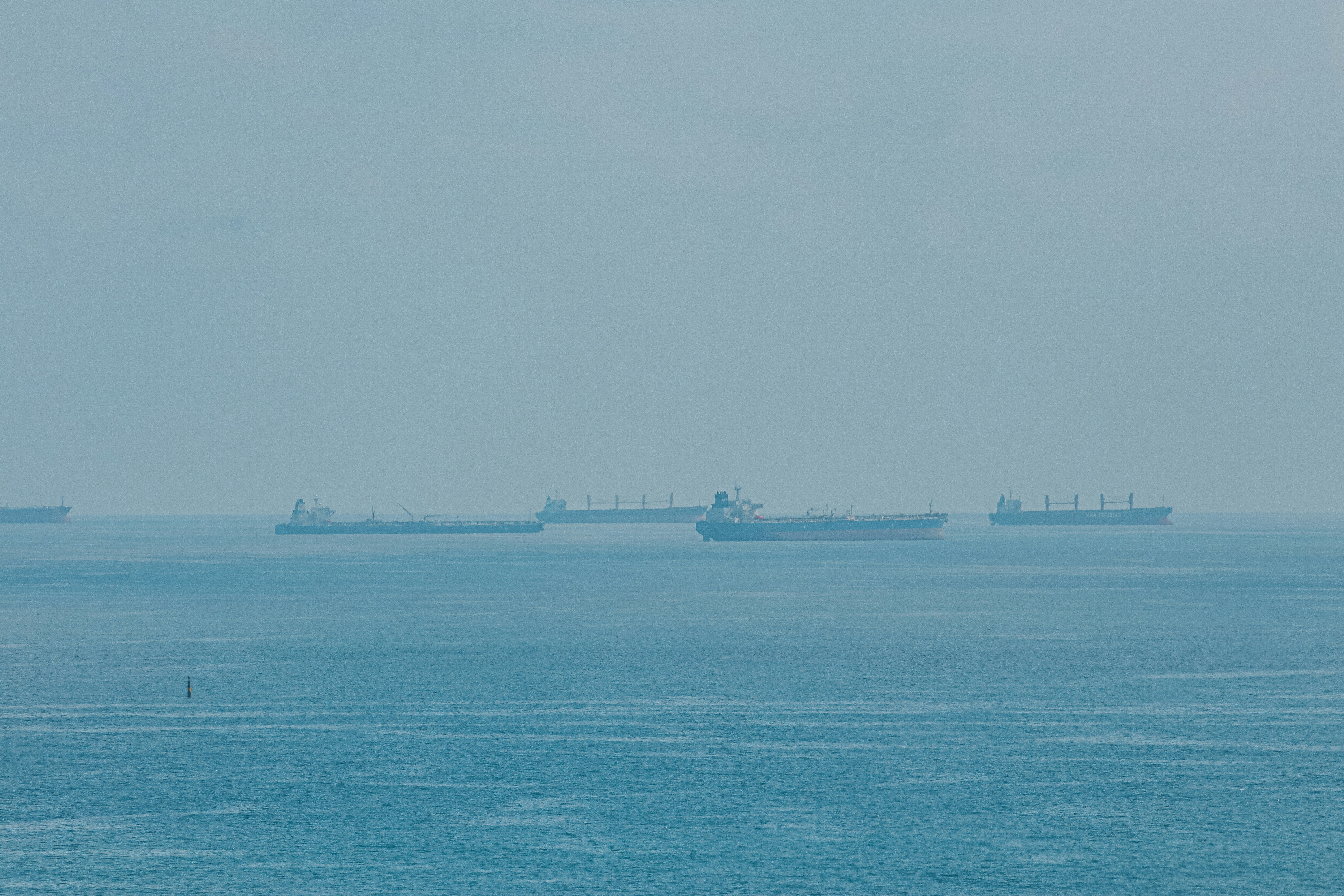 A group of cargo ships anchored in a tranquil sea, shrouded in a soft haze. The calm waters reflect the muted colors of the sky.