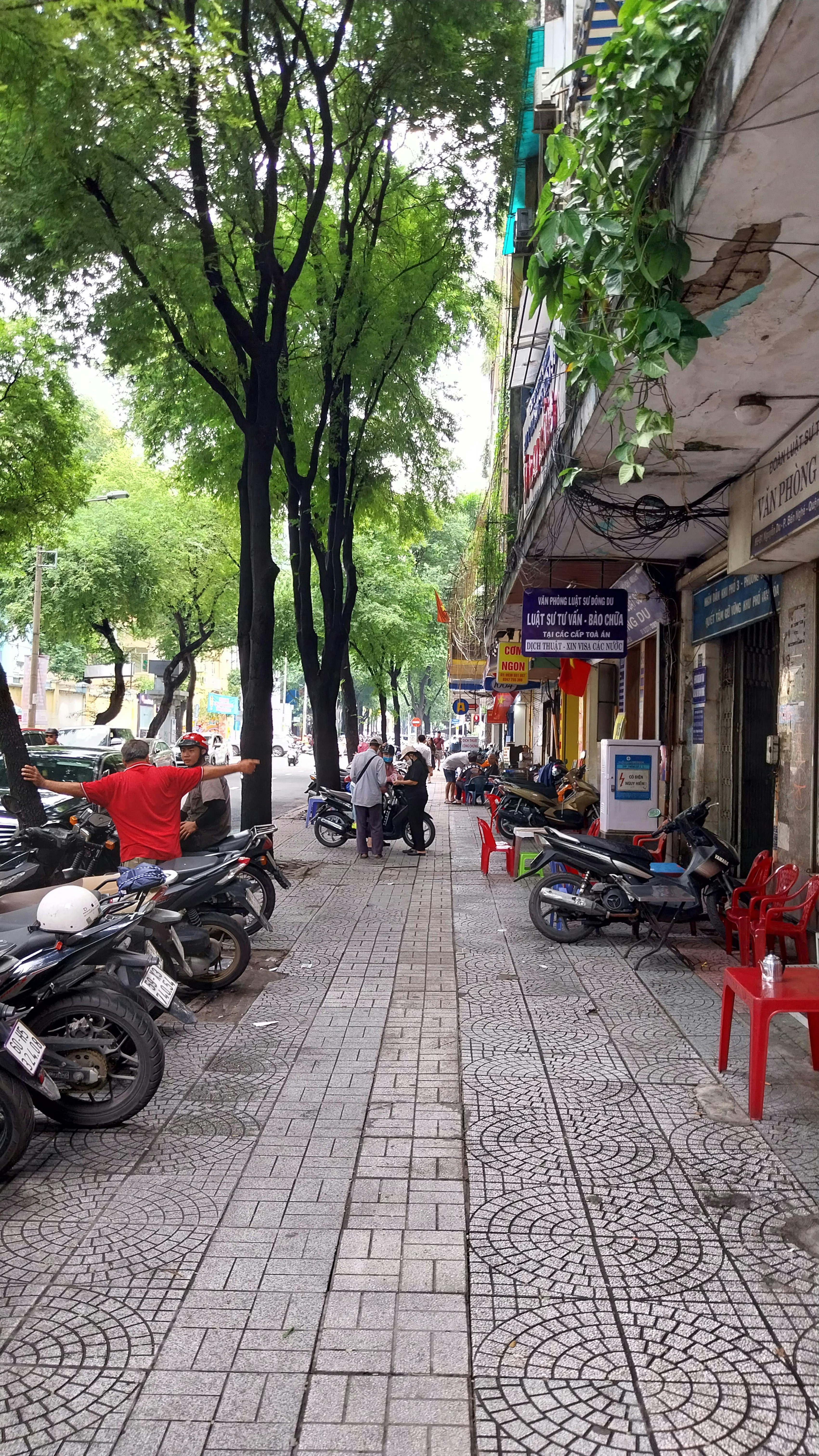 Tree-lined street with parked motorcycles and row of storefronts. A red-shirt figure gestures by the curb, capturing everyday urban life.