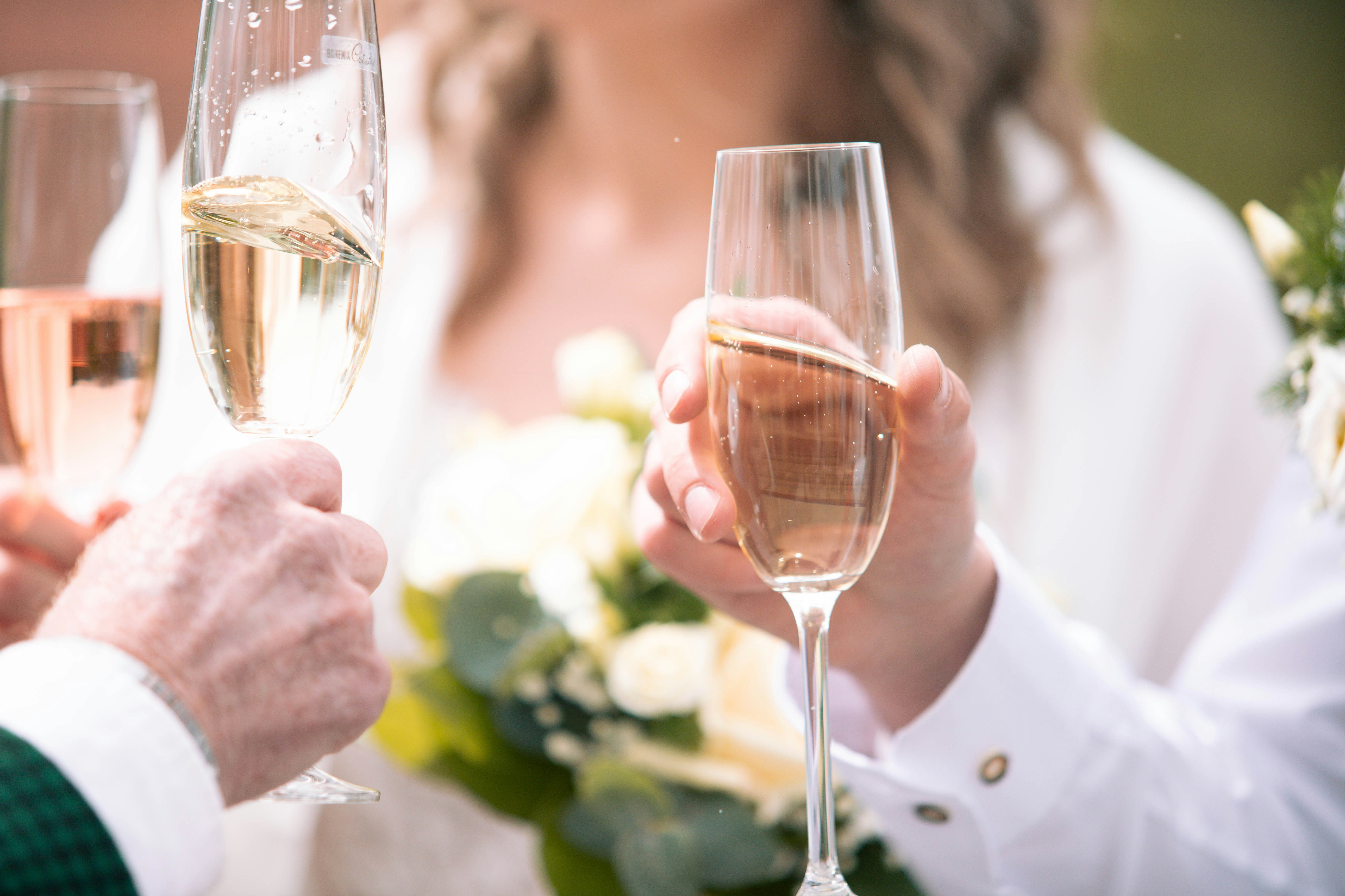 Hands clinking champagne glasses amidst a bouquet of white roses at a celebration.