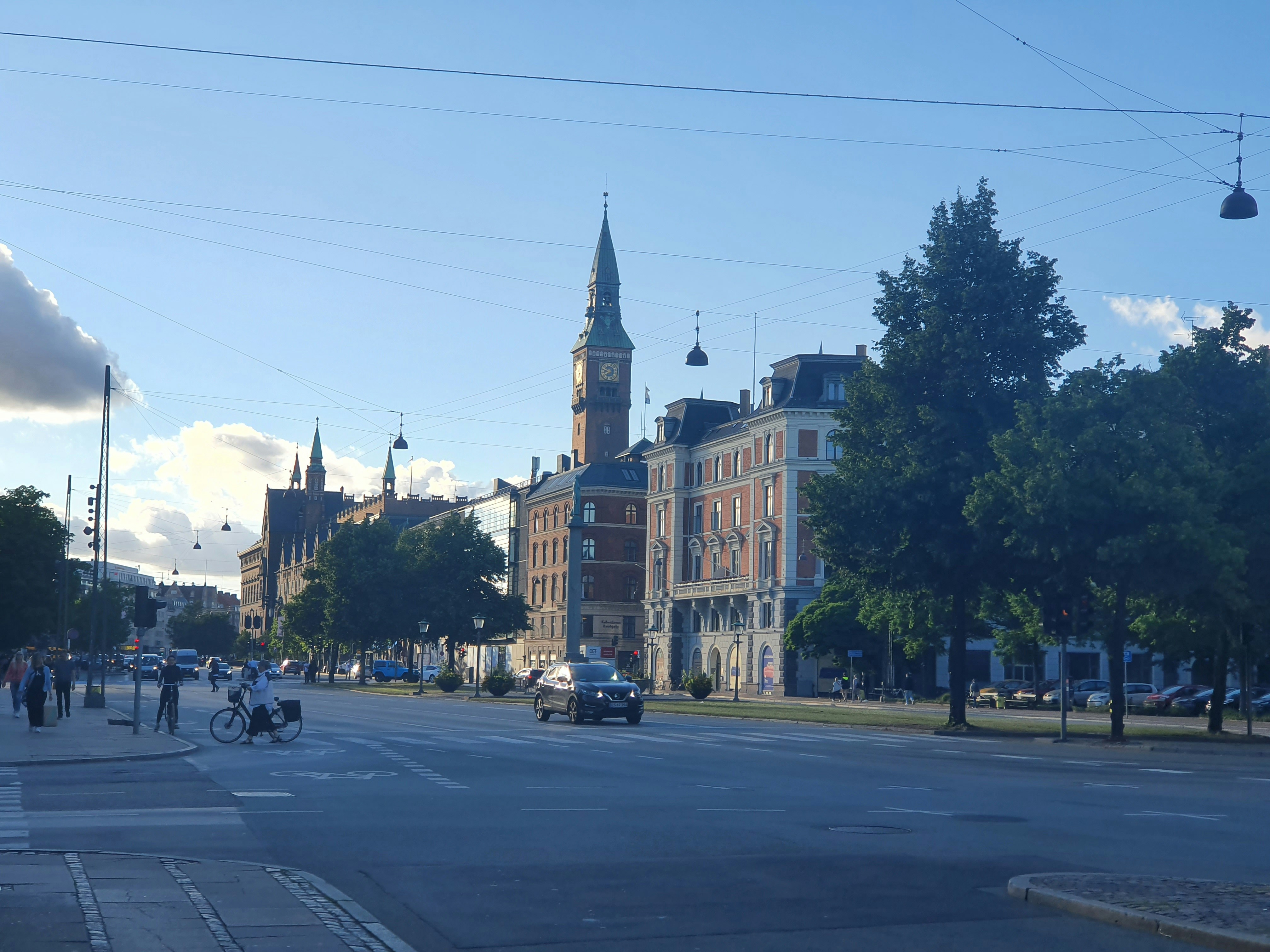 A sunlit city street lined with trees and classic brick buildings leads to a tall church clock tower in the distance.