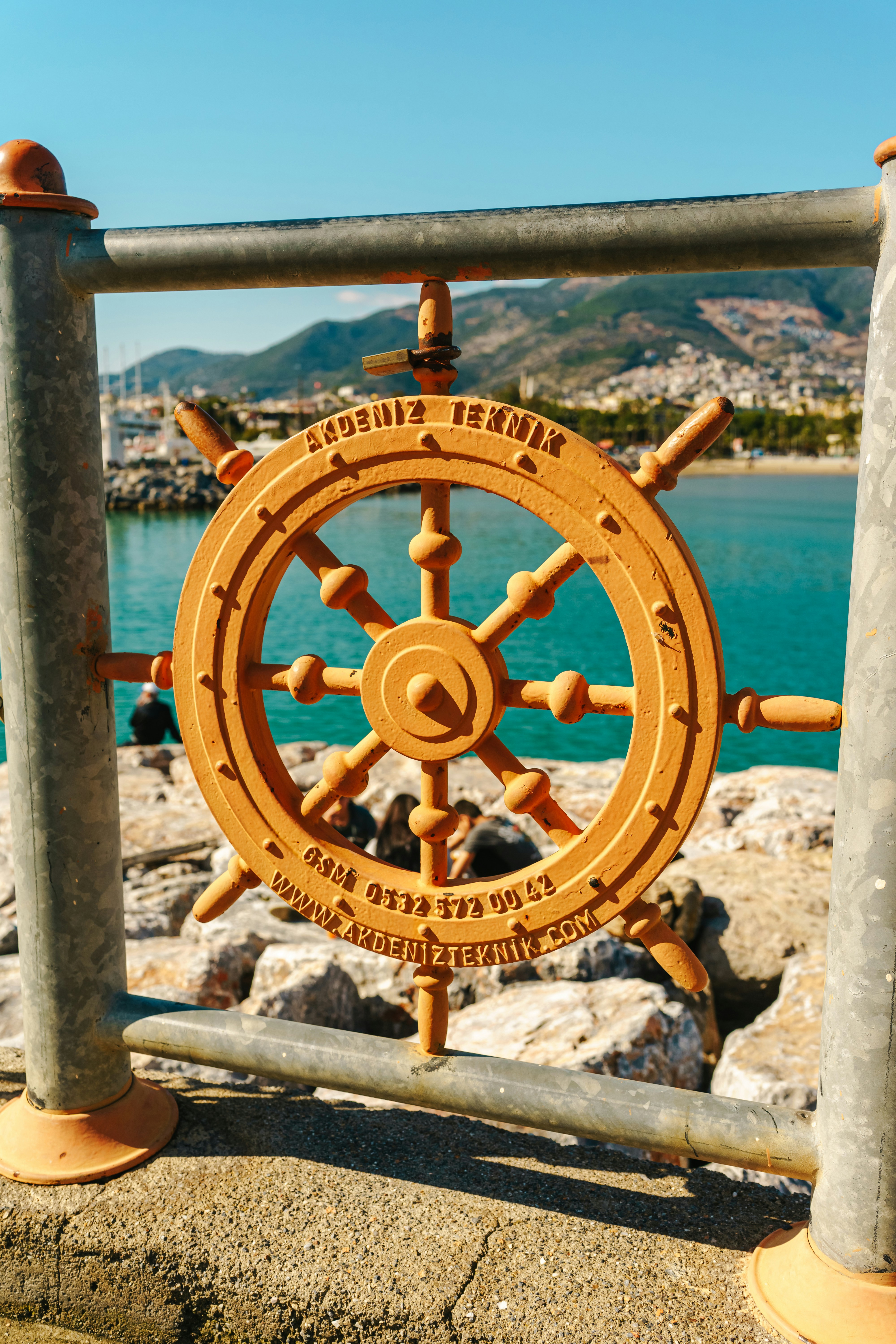 Orange Ship Wheel Framed by Seaside Rail Over Turquoise Harbor