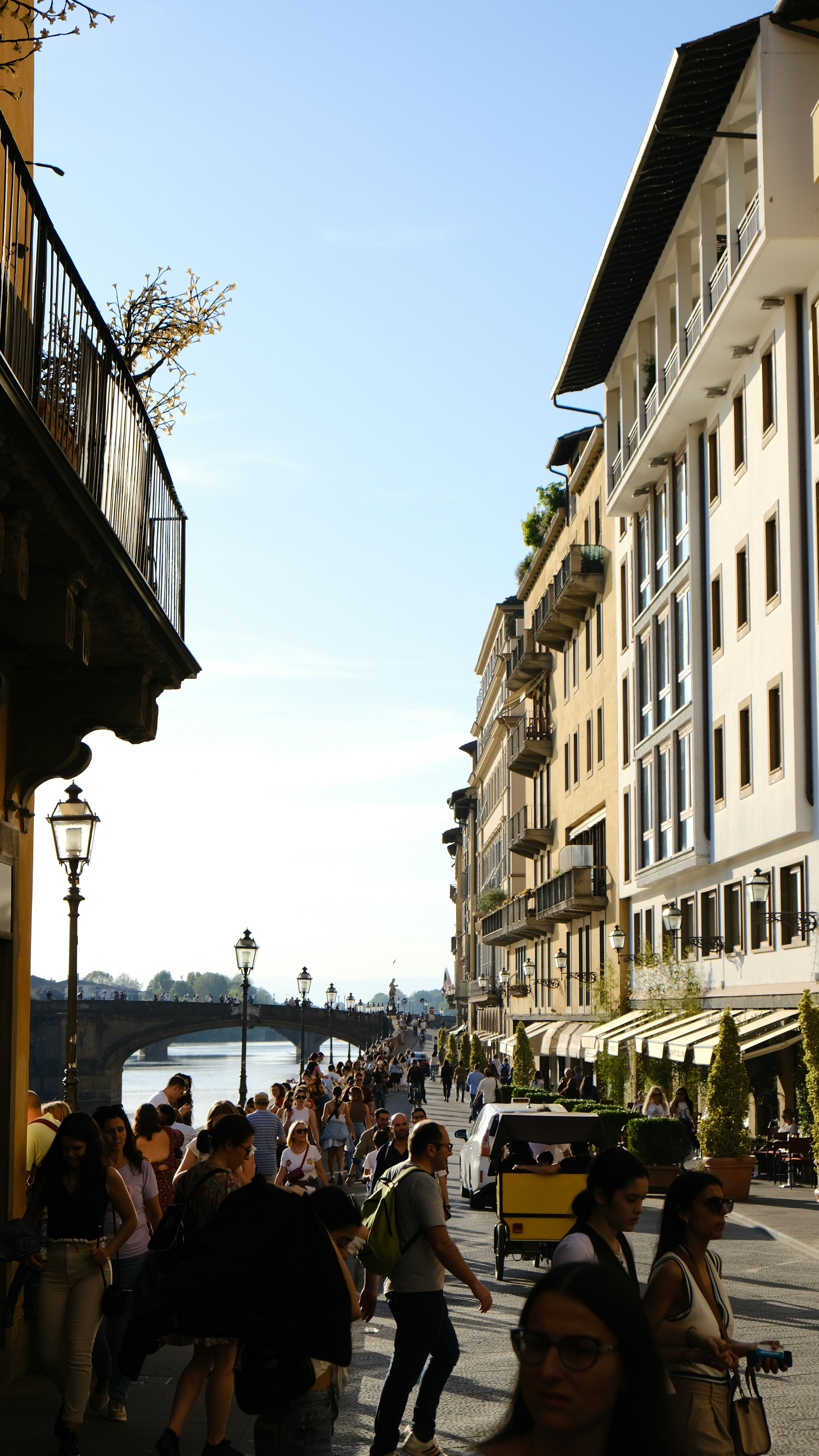 Promenade by the River: Crowds, Arched Bridge, and Sunlit Facades