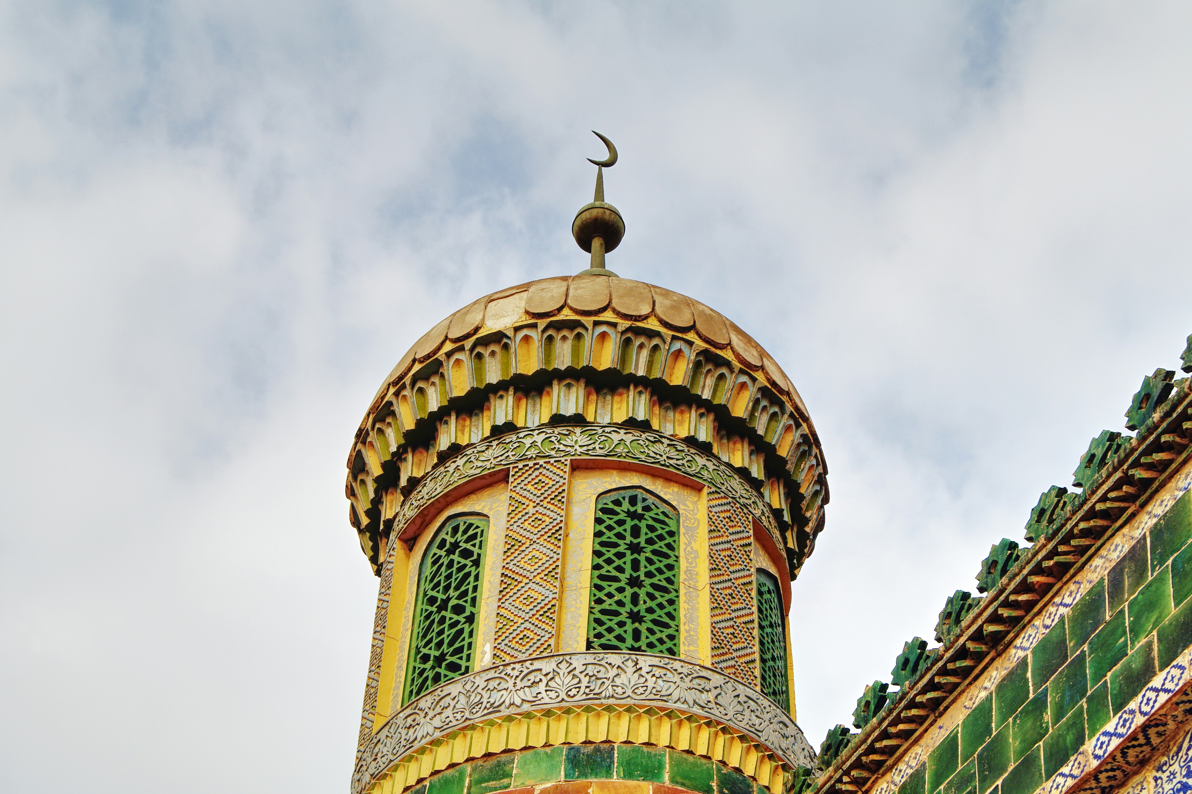 Close-up photograph of a mosque minaret dome featuring green lattice windows and ornate gold-green tilework beneath a crescent finial against a pale blue sky.