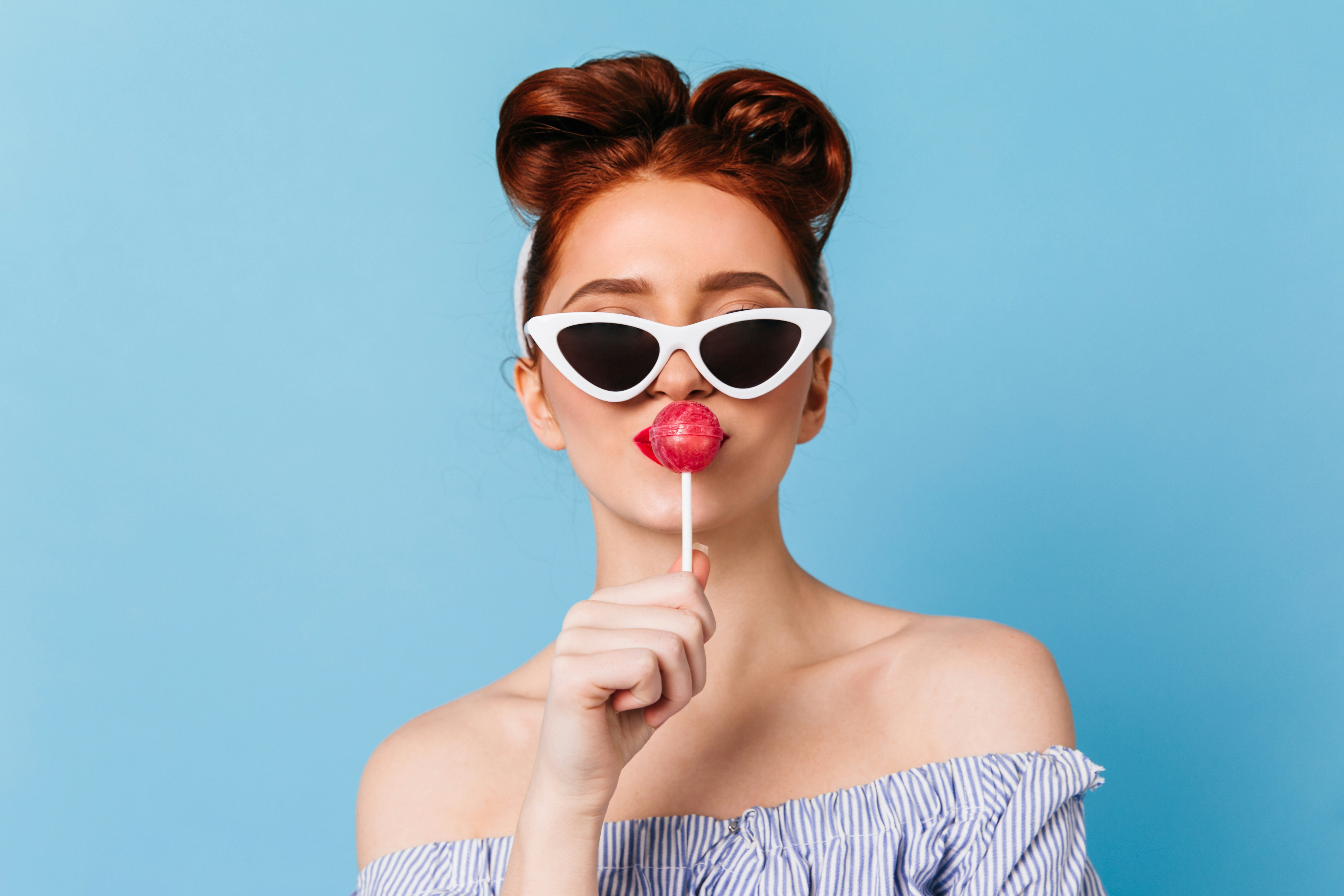 Young woman in stylish white sunglasses enjoying a lollipop against a vibrant blue background.