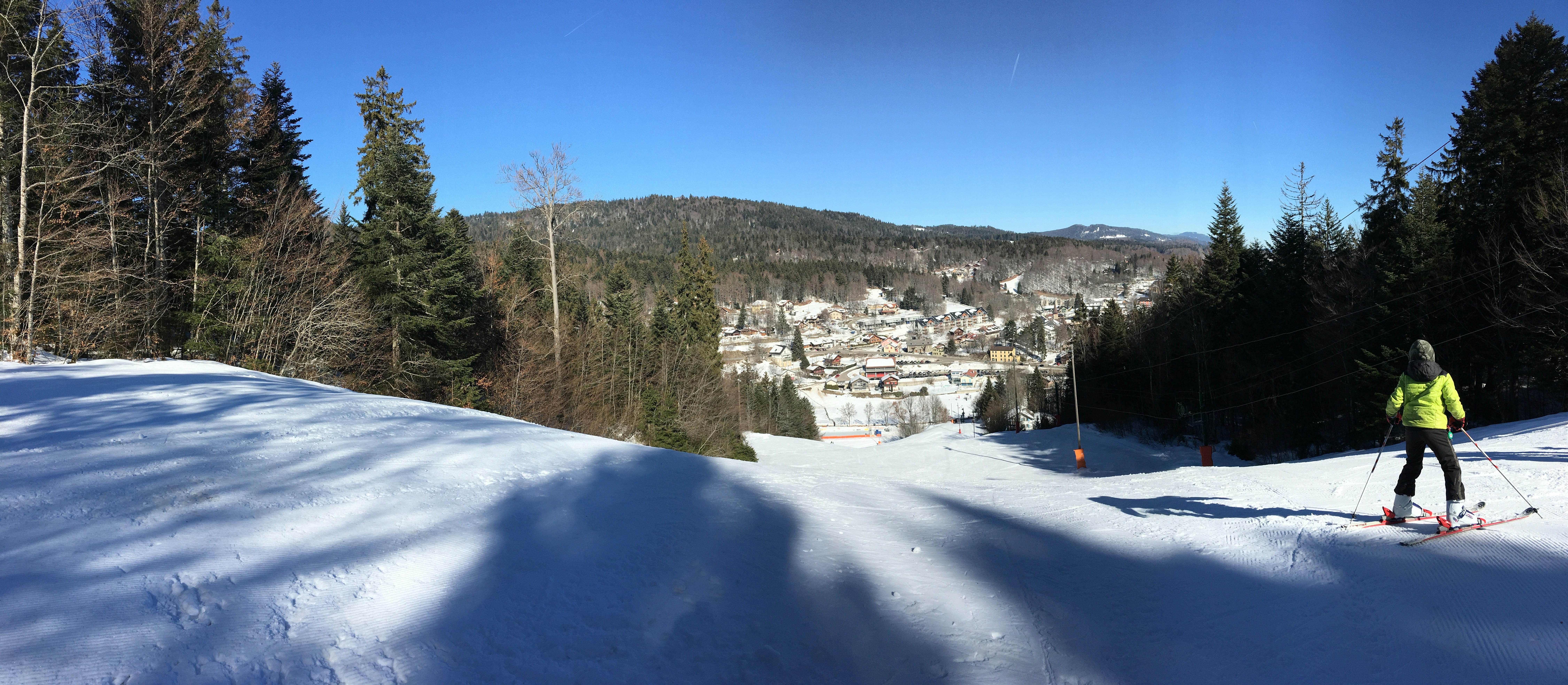 Skier glides along a sunlit groomed slope as a distant alpine town rises beyond evergreen trees under a clear blue sky.