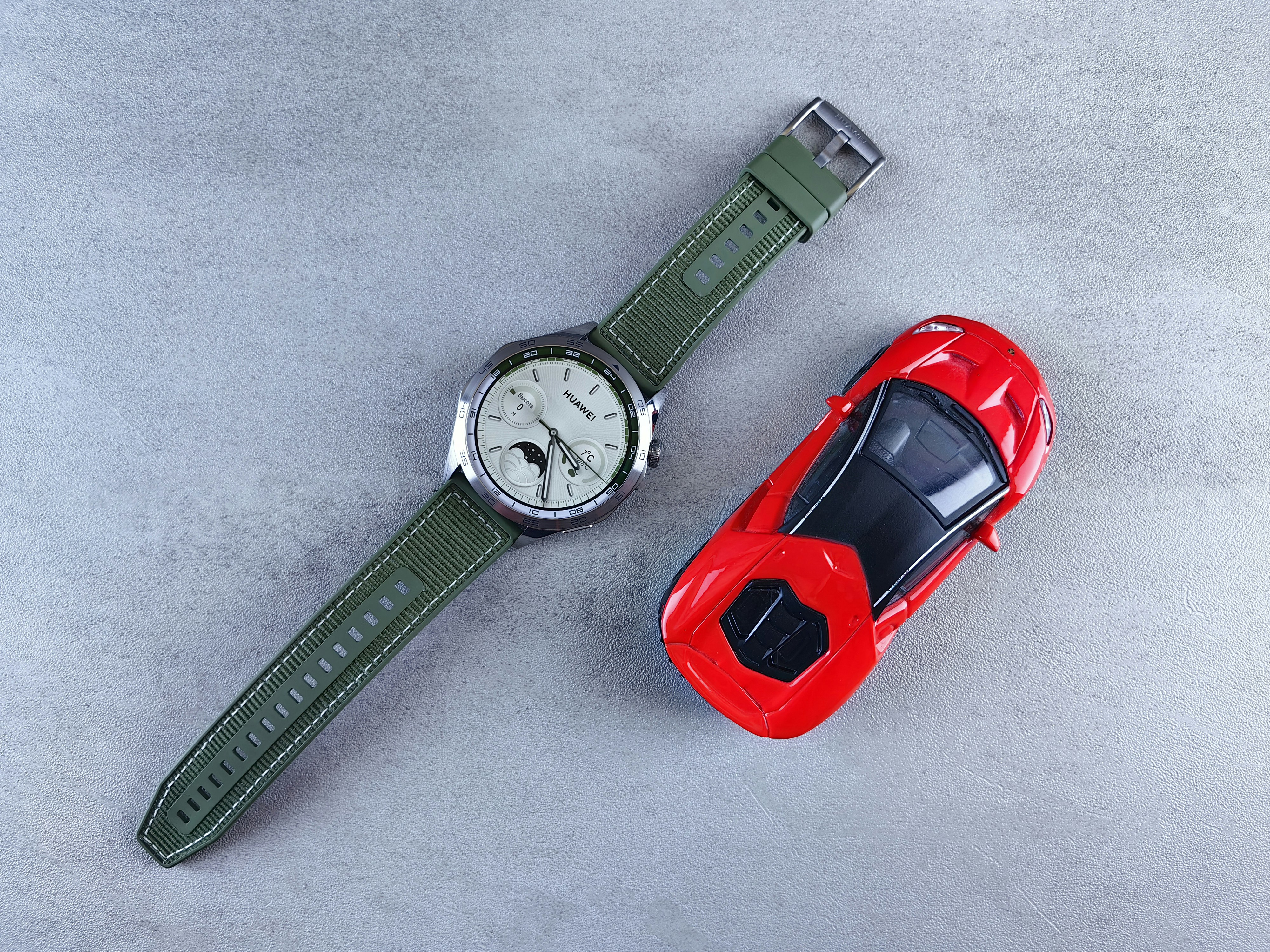 Photograph of a green-strap watch beside a red toy sports car on a gray textured surface.