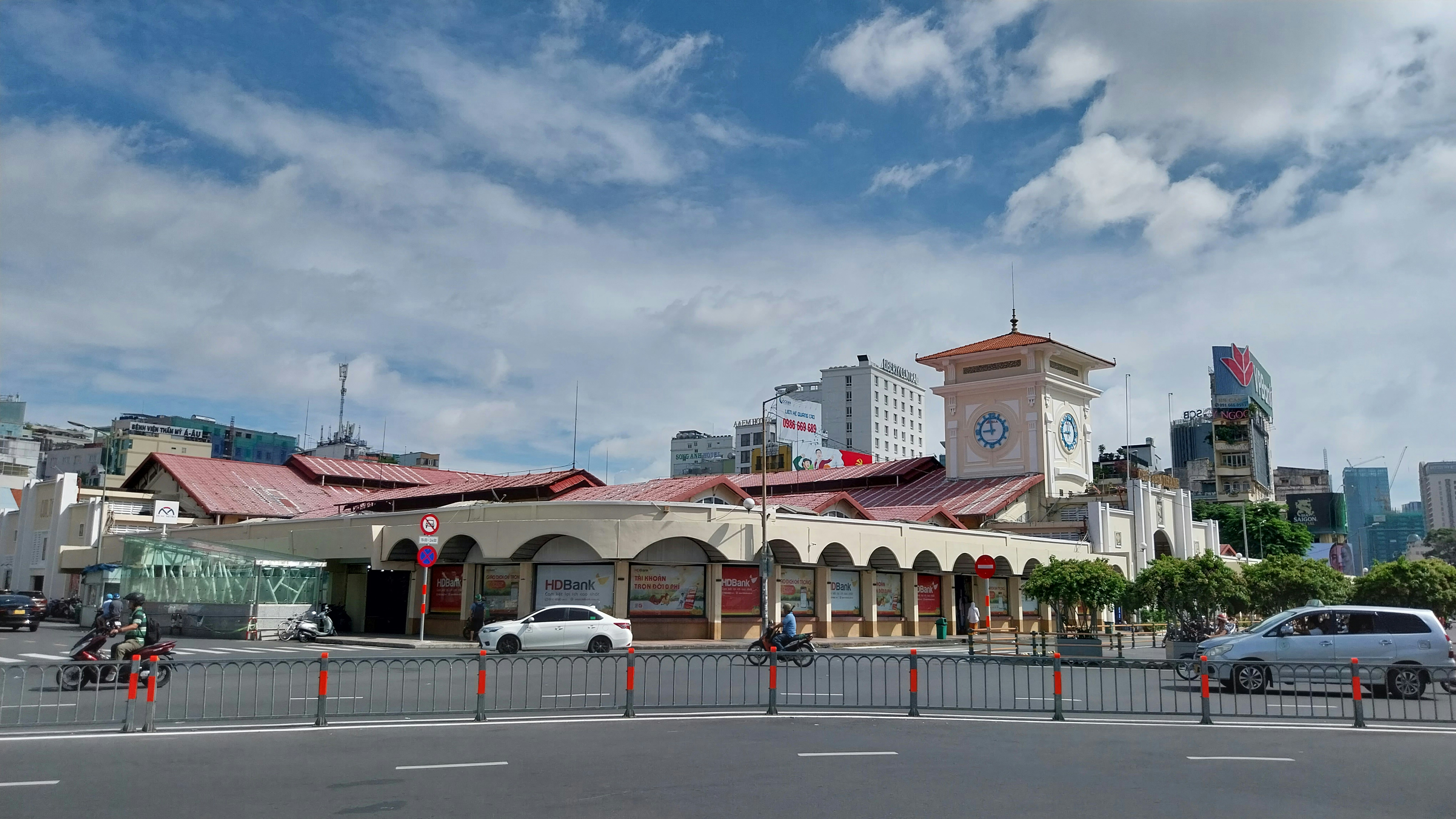 Clock-topped transit station along a wide avenue, with vehicles and pedestrians in motion and a blue sky above. Painting-like façade details and a row of arched storefronts anchor the scene.