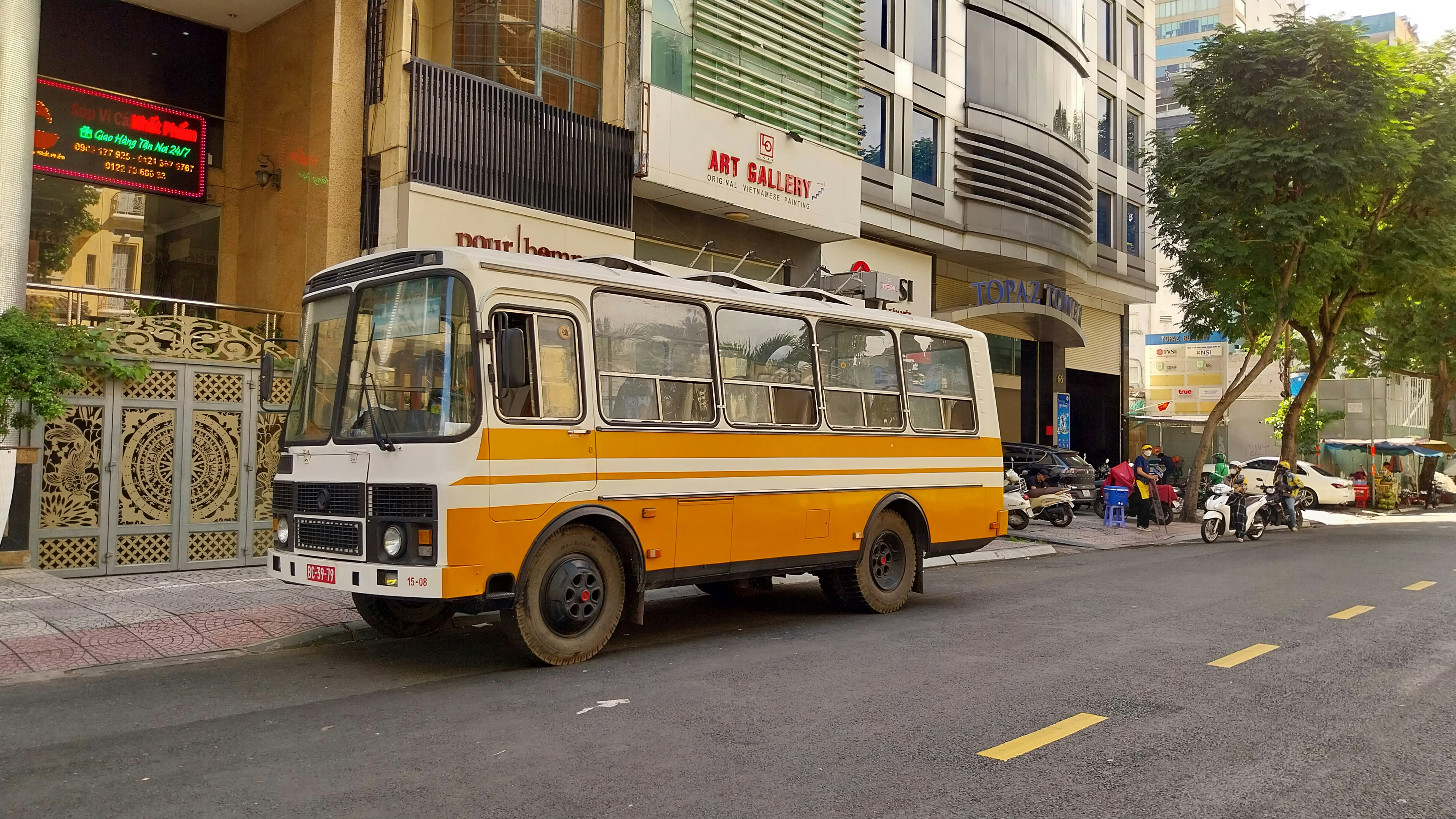A vintage yellow bus parked on a bustling city street, surrounded by modern architecture and greenery.