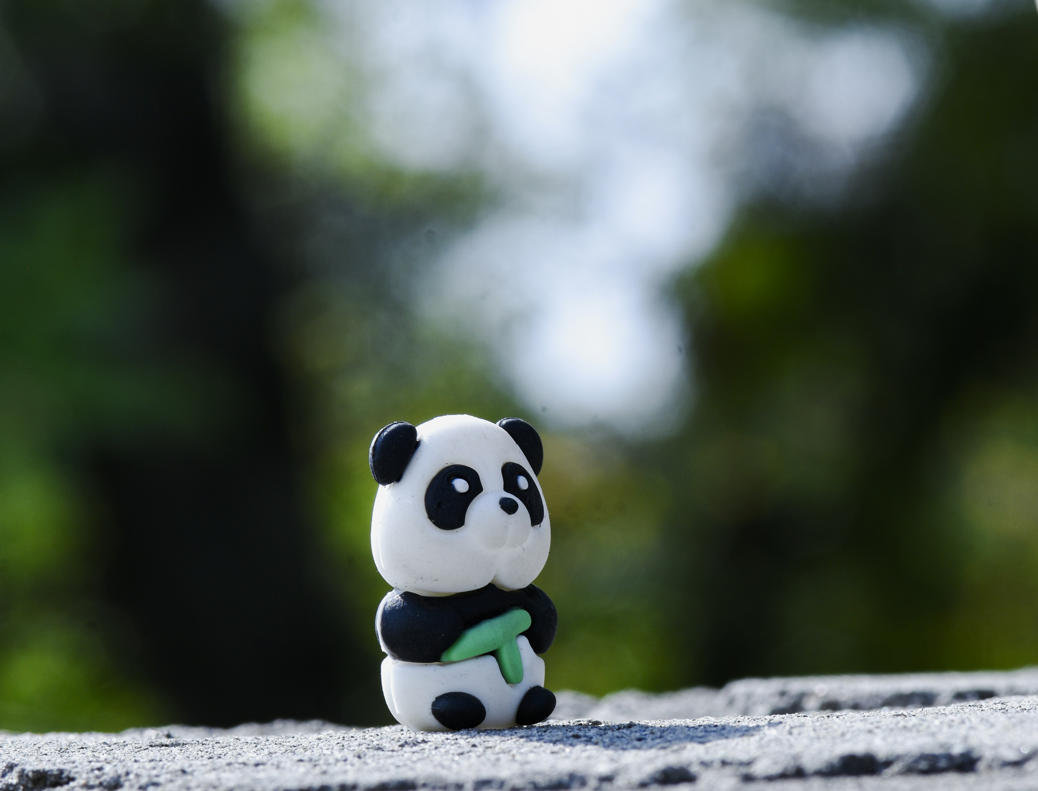 A charming panda figurine holding a green bamboo stick, set against a softly blurred natural background. The playful design contrasts with the serene environment.