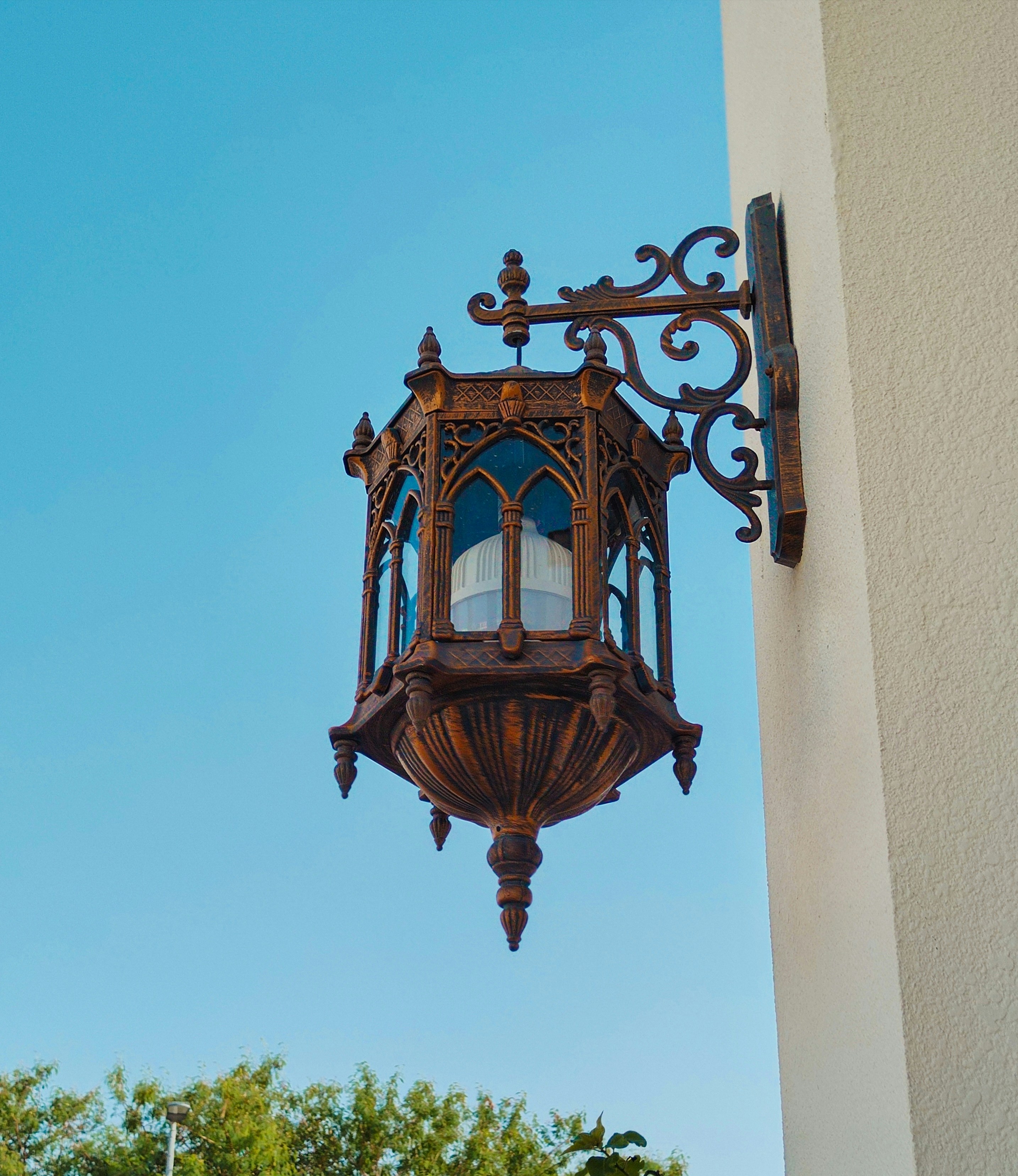 Ornate iron lantern mounted on a pale wall against a clear blue sky. The intricate metalwork and glass enclosure are the focal point of this outdoor composition.