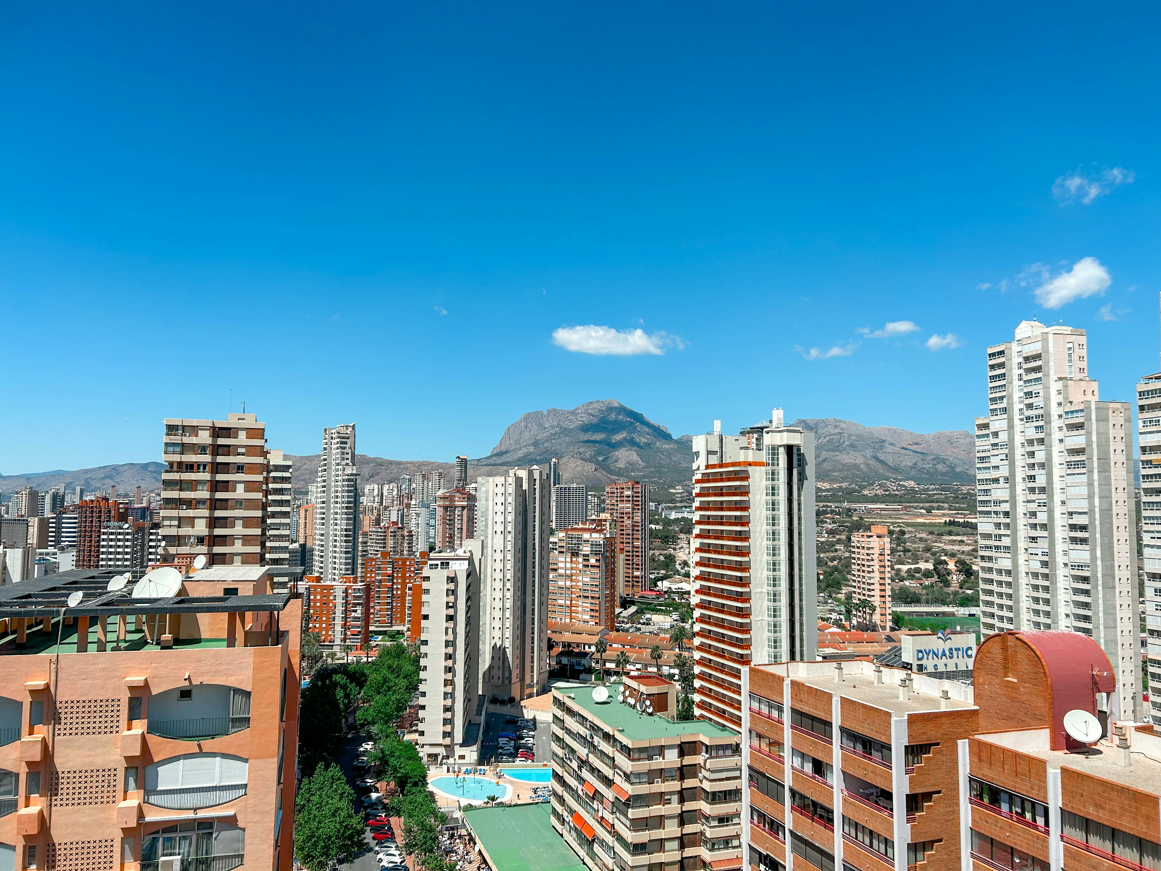 A panoramic view of a bustling cityscape featuring high-rise buildings and a distant mountain backdrop. The scene is framed by vibrant blue skies.