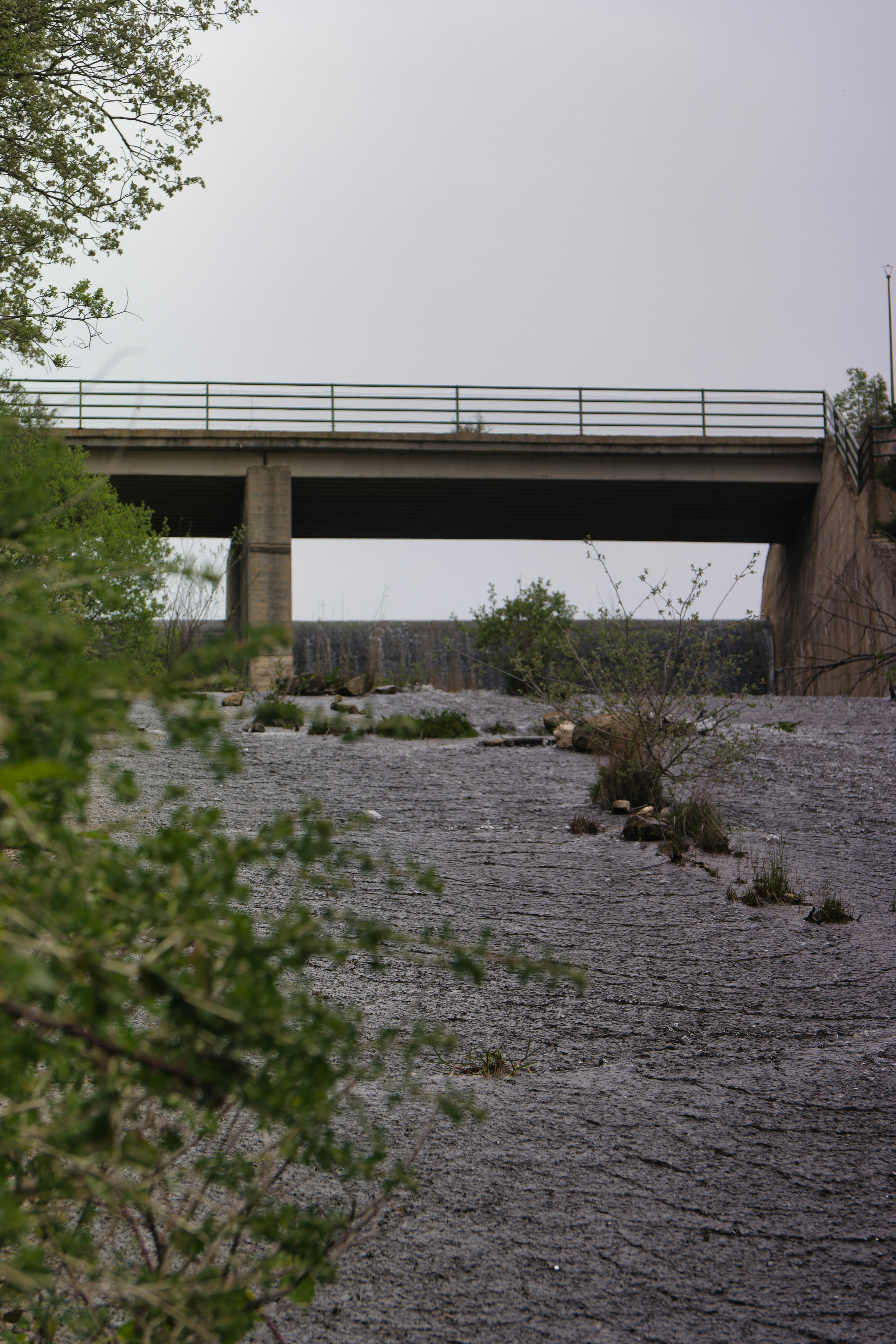 A photograph of a concrete bridge spanning a shallow, rocky riverbed. Greenery frames the foreground as a metal railing runs along the bridge beneath a gray, overcast sky.