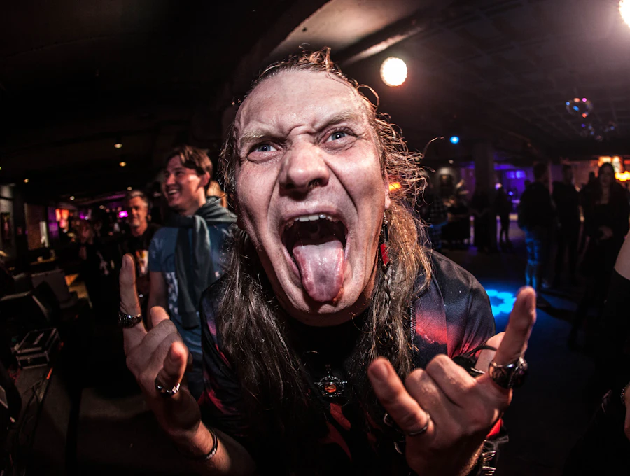 A long-haired man leans toward the lens with his tongue out and hands raised in a rock gesture inside a dim club. Colored lights and other crowd members blur in the background.