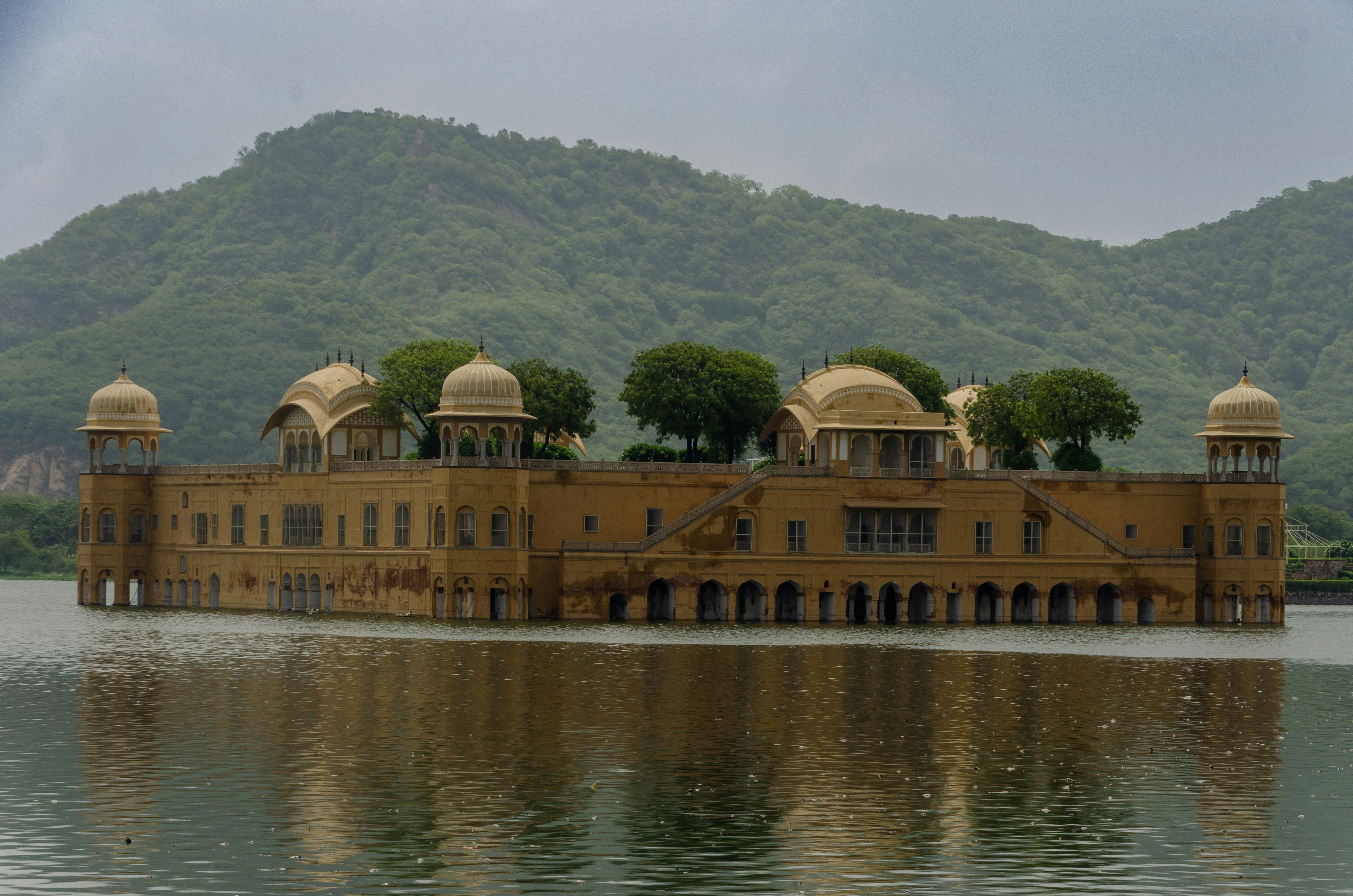 Majestic palace surrounded by water, set against a backdrop of lush green hills. The structure features domed roofs and is partially submerged, creating a serene reflection on the water's surface.