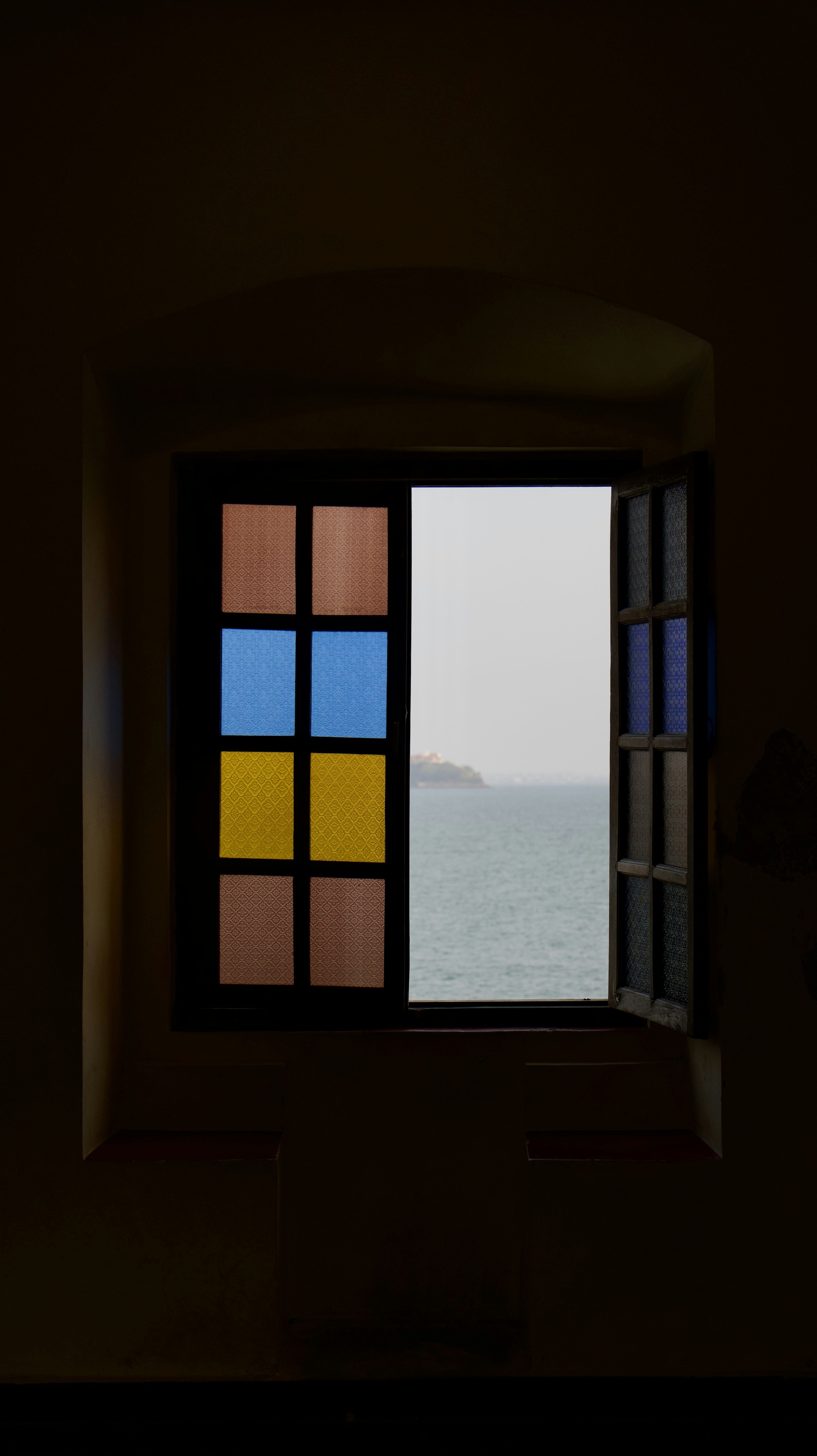 Dim interior with a left panel of colored glass. This photograph highlights light filtering through stained panes as the open right pane reveals the sea.