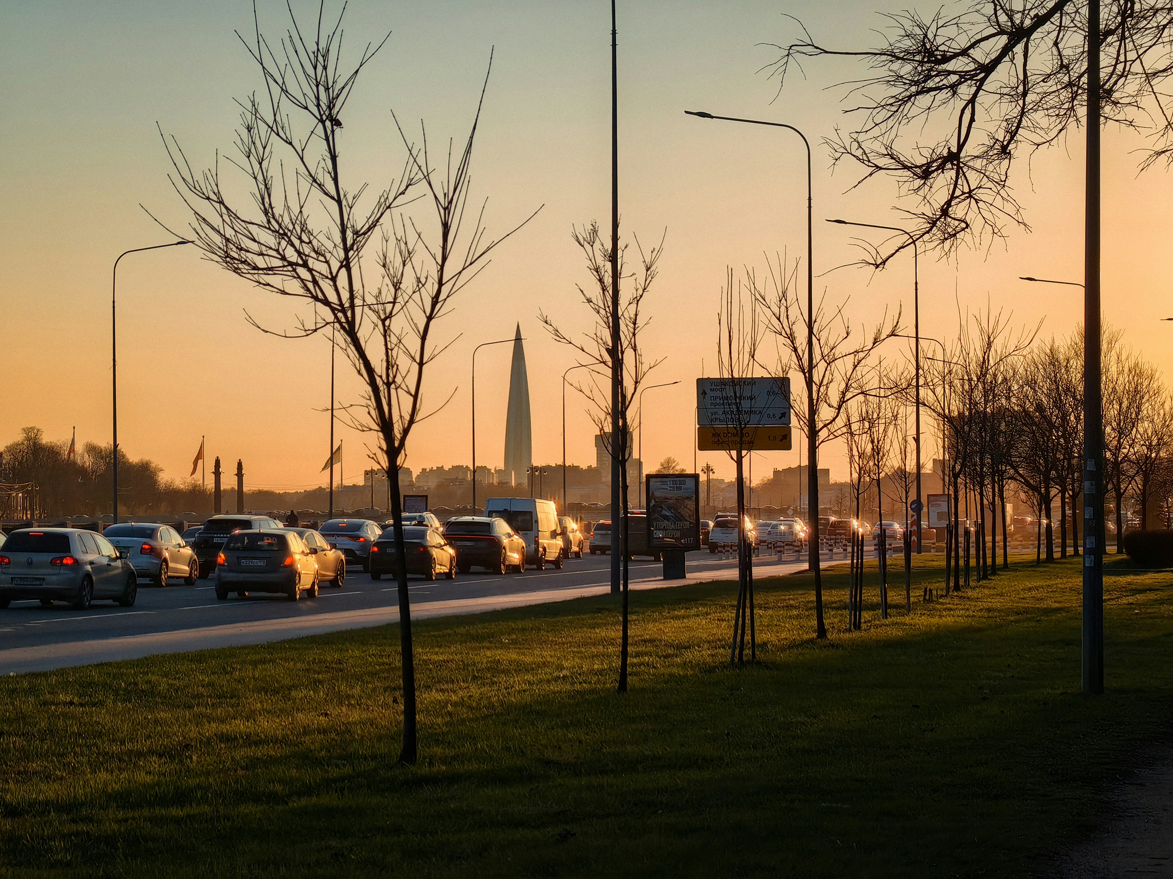 Golden-hour street scene with a row of bare trees and cars along a traffic-filled boulevard. A distant obelisk silhouette rises against a warm sunset.