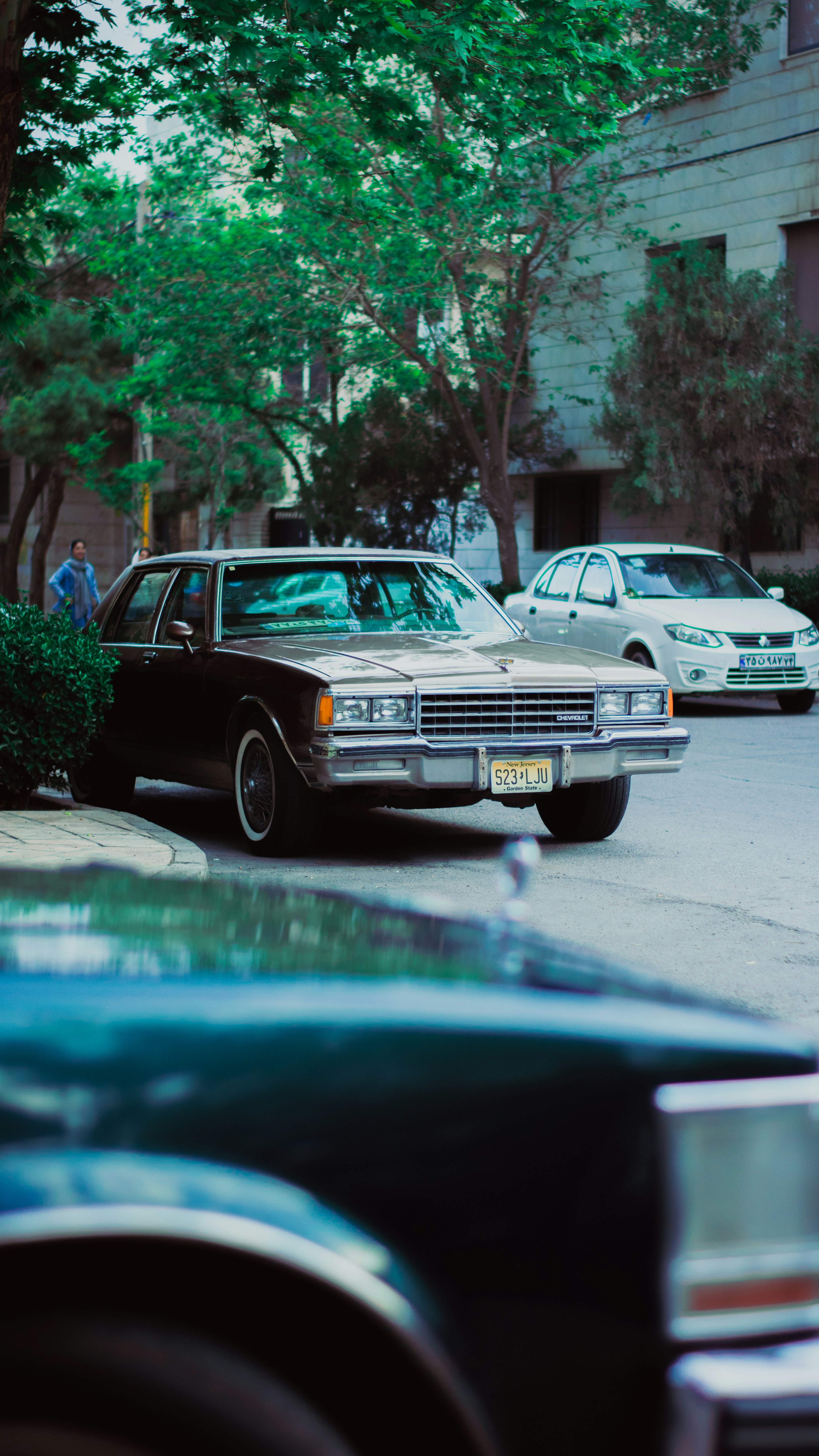 Classic car parked on an urban street with trees and modern vehicles in the background.