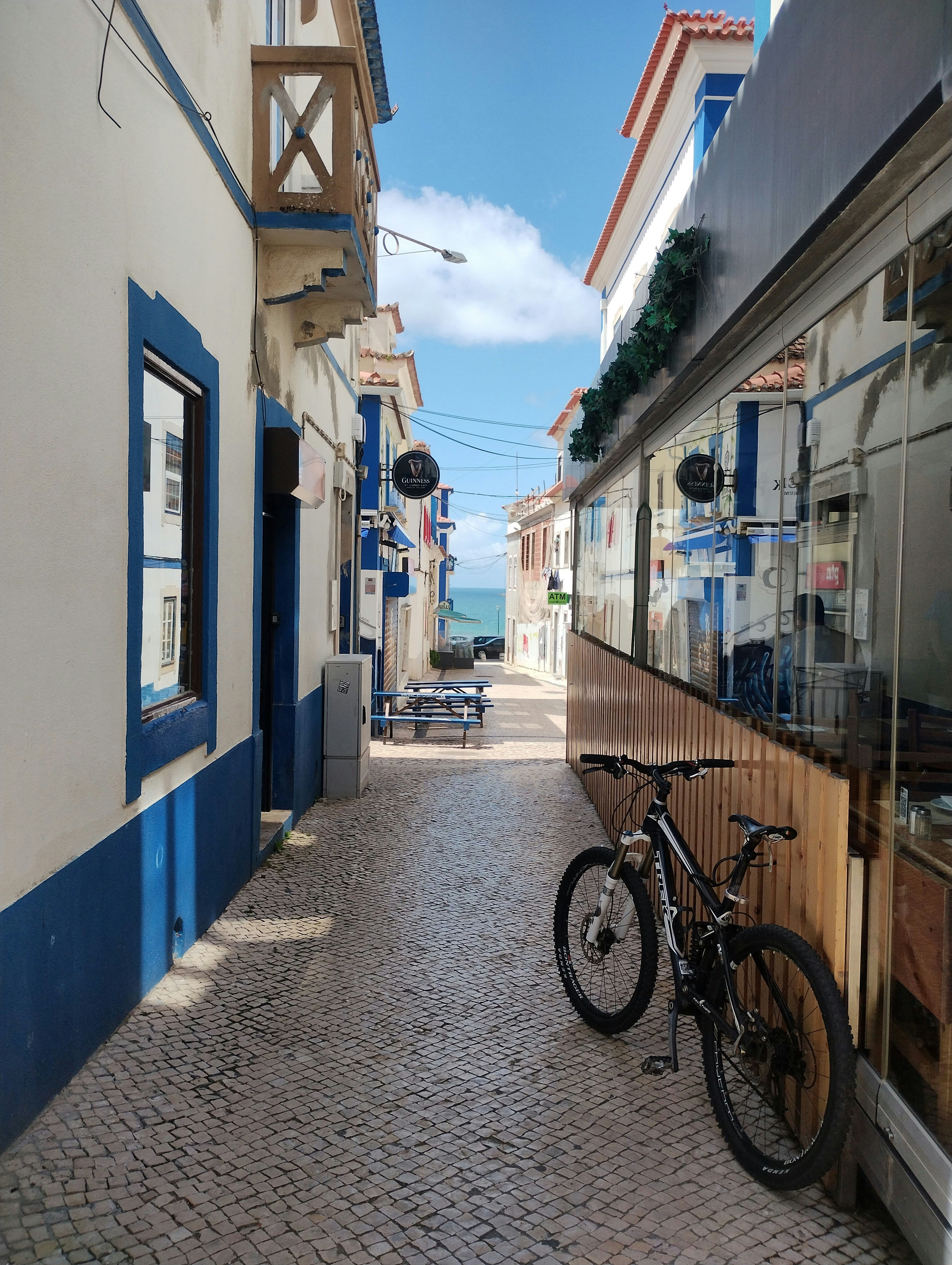 Sunlit alley framed by blue-and-white shutters leads toward a distant sea. A bicycle rests beside a glass storefront on a cobblestone path.
