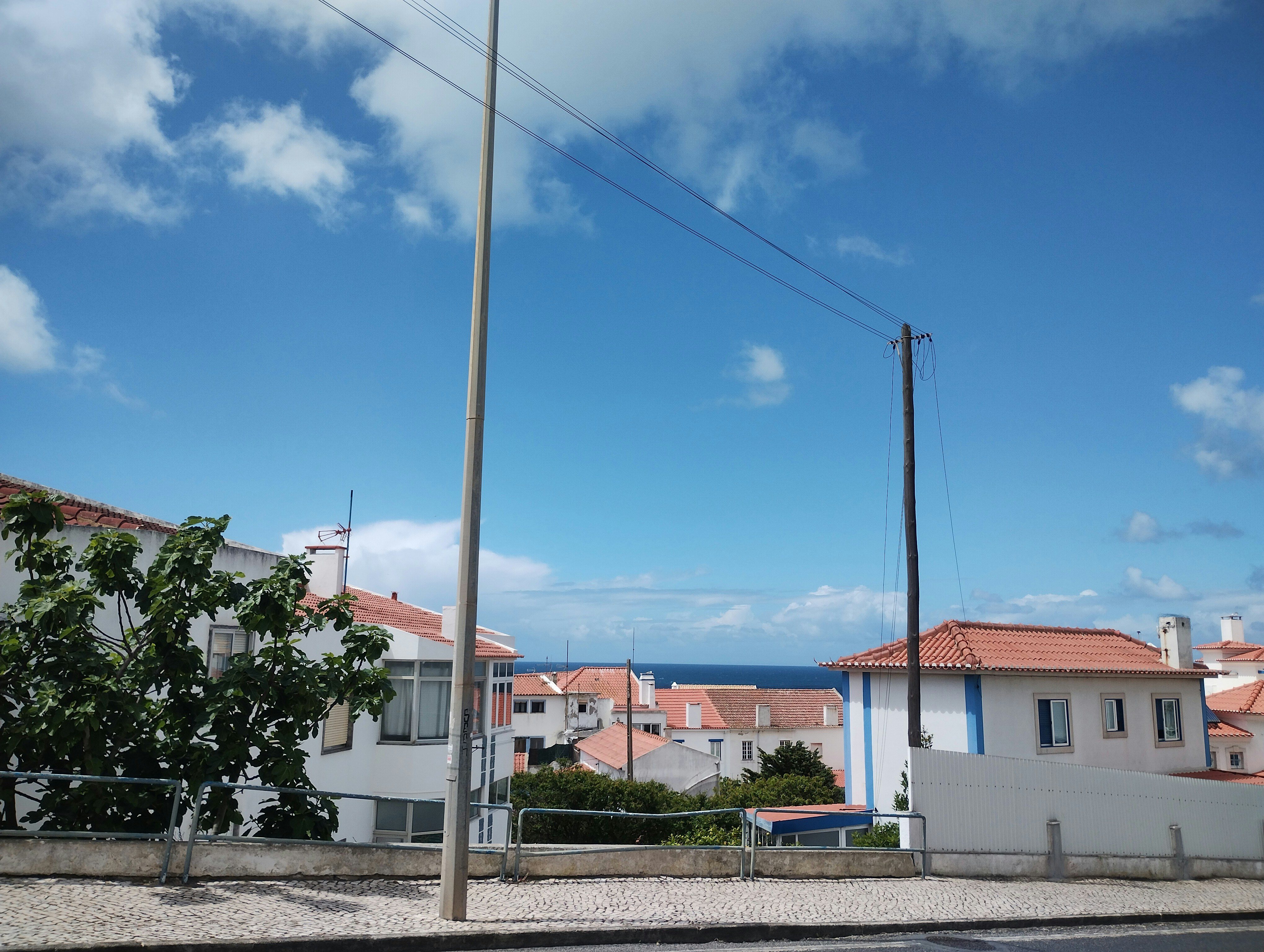Coastal street scene with white houses and red-tiled roofs under a clear blue sky; distant sea on the horizon.