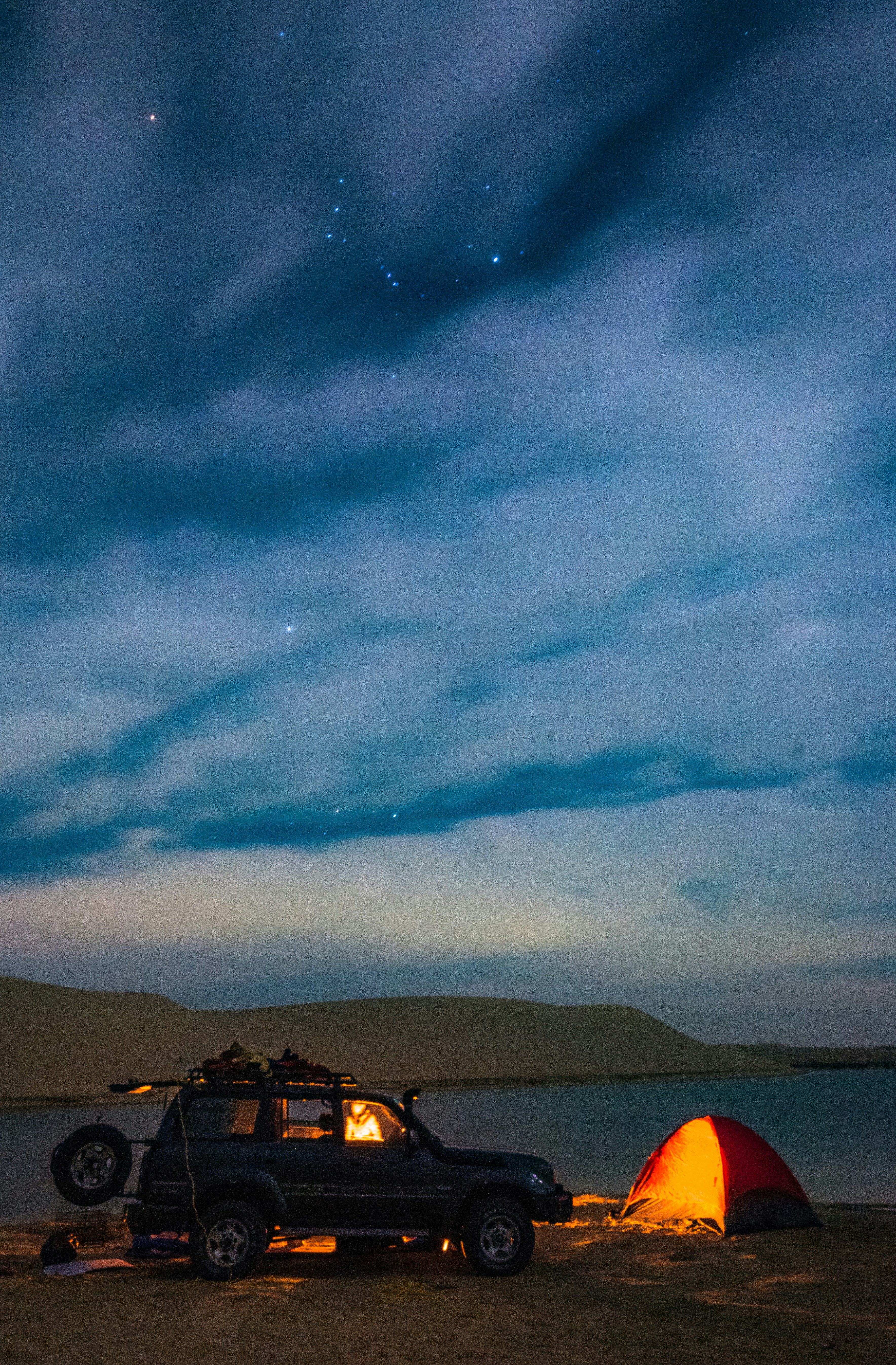 A nocturnal photograph of a rugged SUV with gear on a lakeshore, lit by a warm campfire beside a glowing red-orange tent. A starry, cloud-swept sky arches over the calm water.