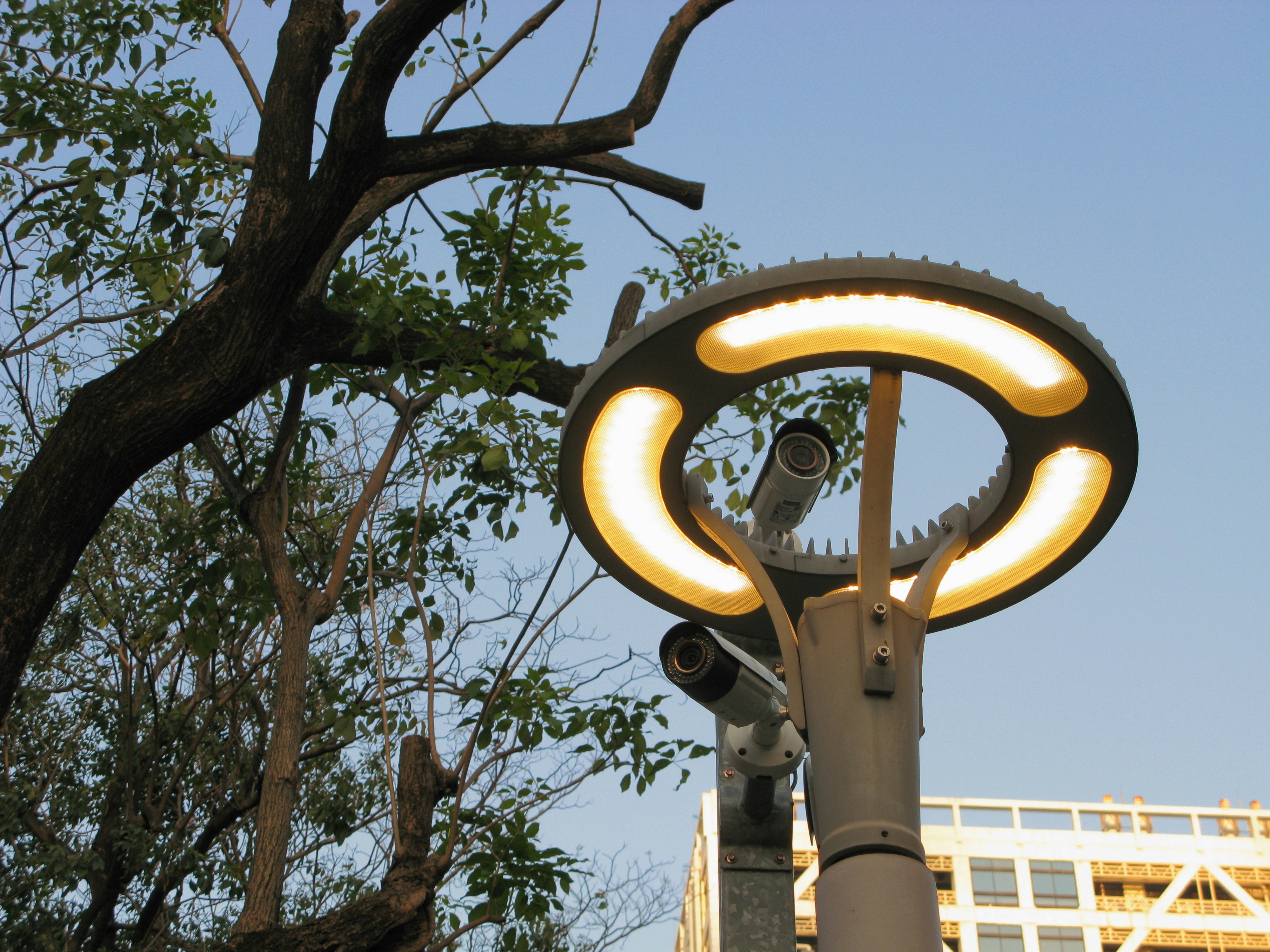 Circular LED ring streetlight with two cameras rises against a clear blue sky. A modern building and tree line form the backdrop.