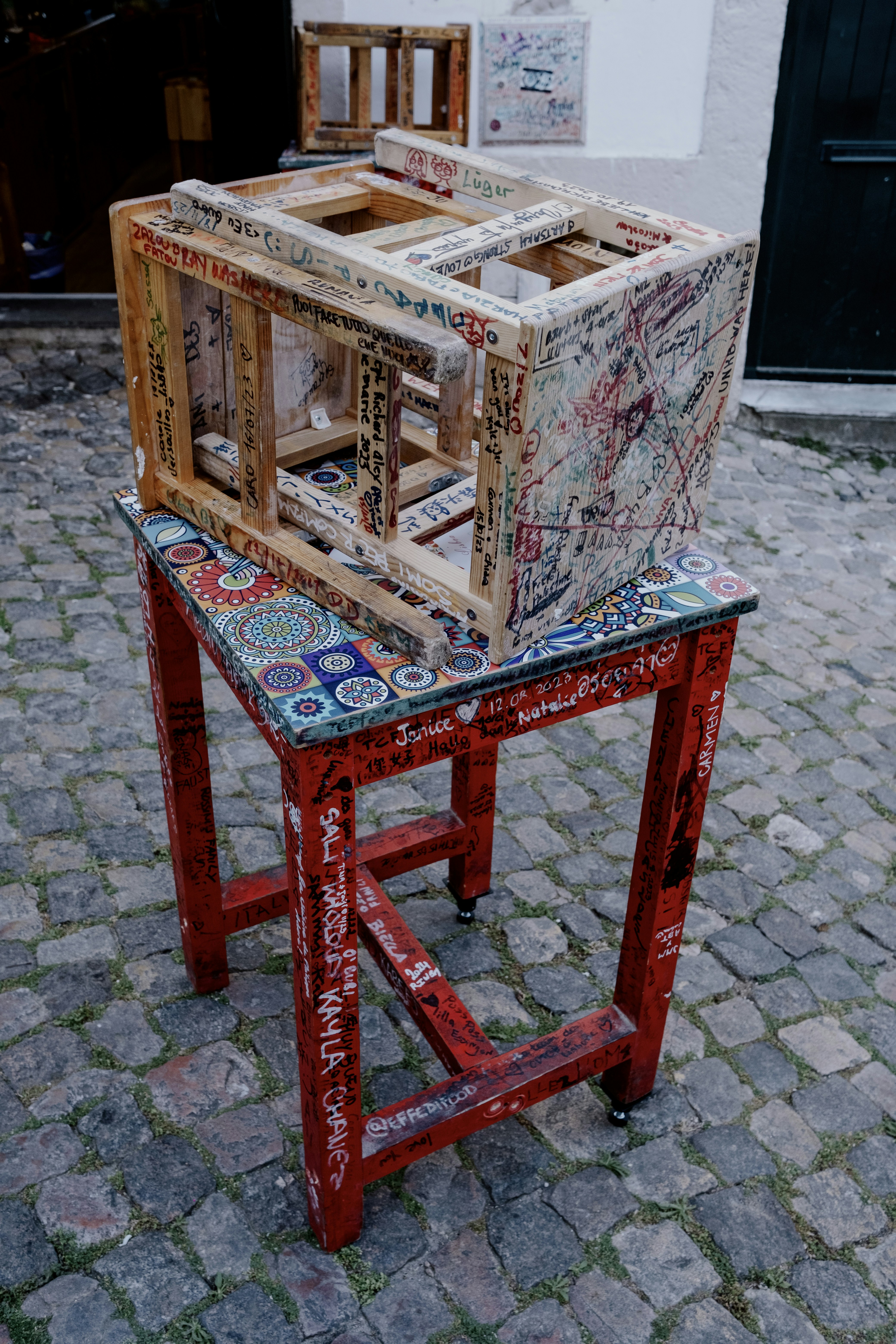 Graffiti-Adorned Crate Balancing on a Mosaic Stool in a Cobblestone Alley