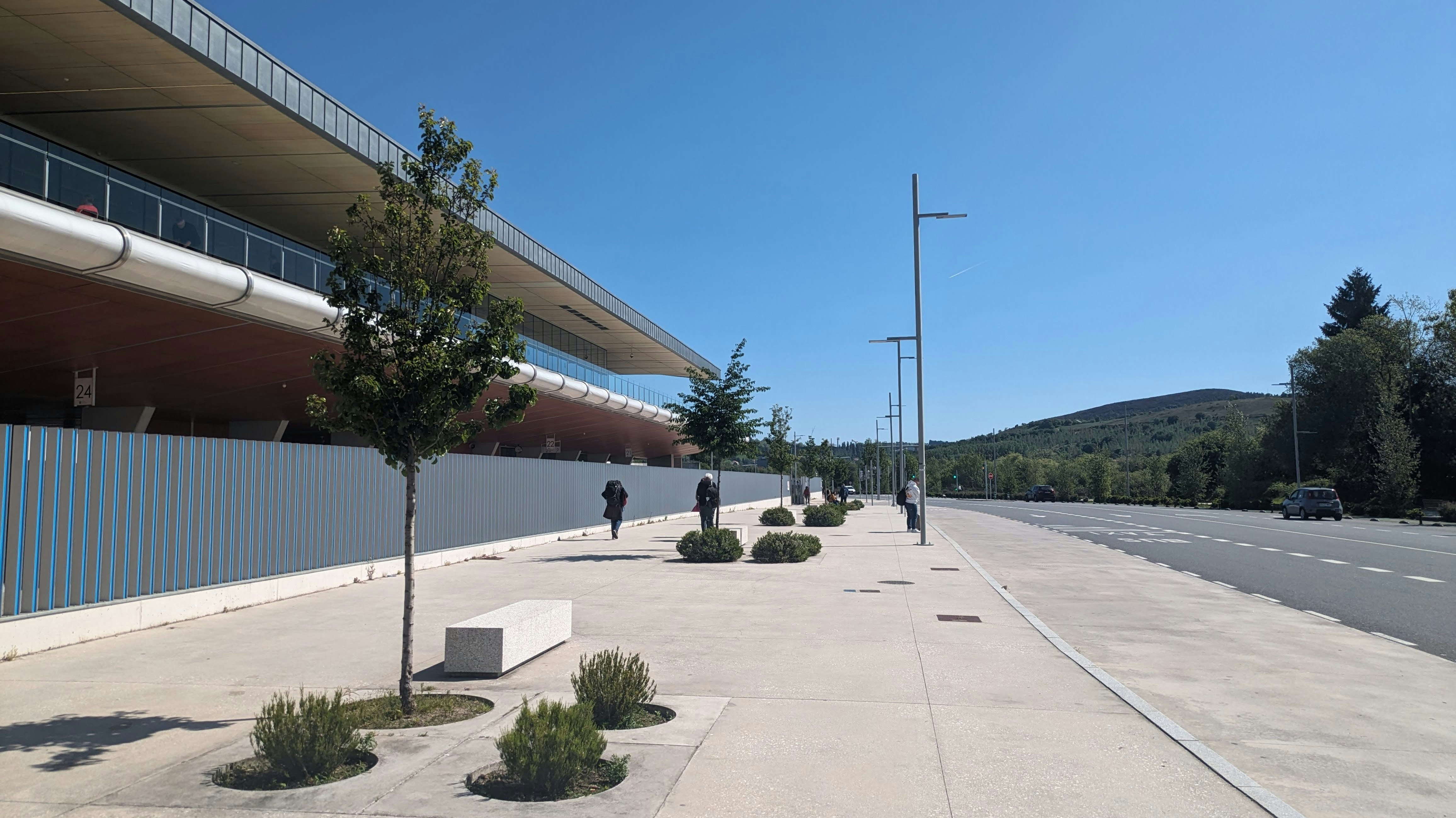 Wide sidewalk beside a modern cantilevered building as cyclists and pedestrians move along the boulevard beneath a bright blue sky.
