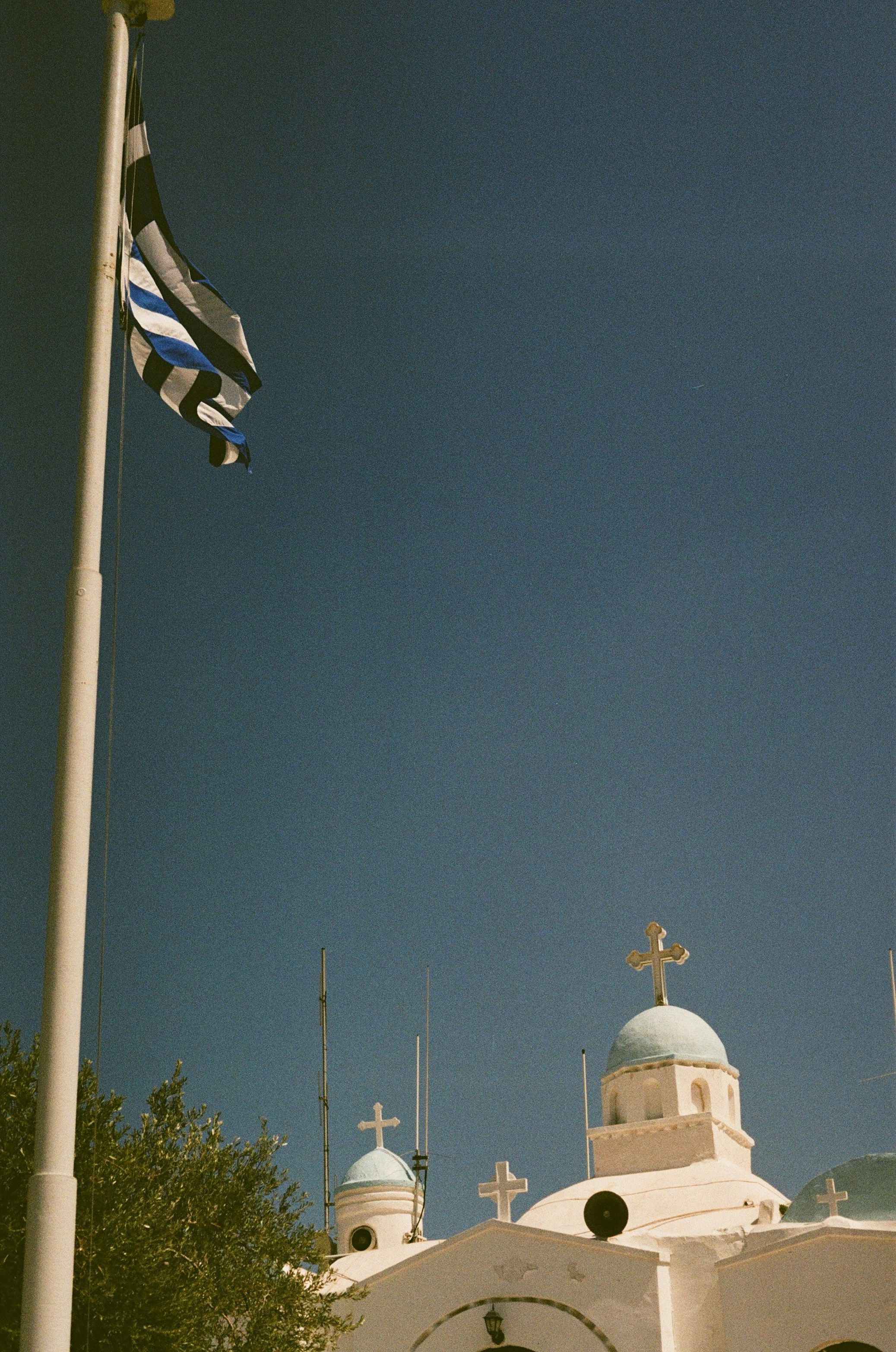 Flagpole on the left frames a sunlit Orthodox church with white domes and crosses against a clear blue sky.