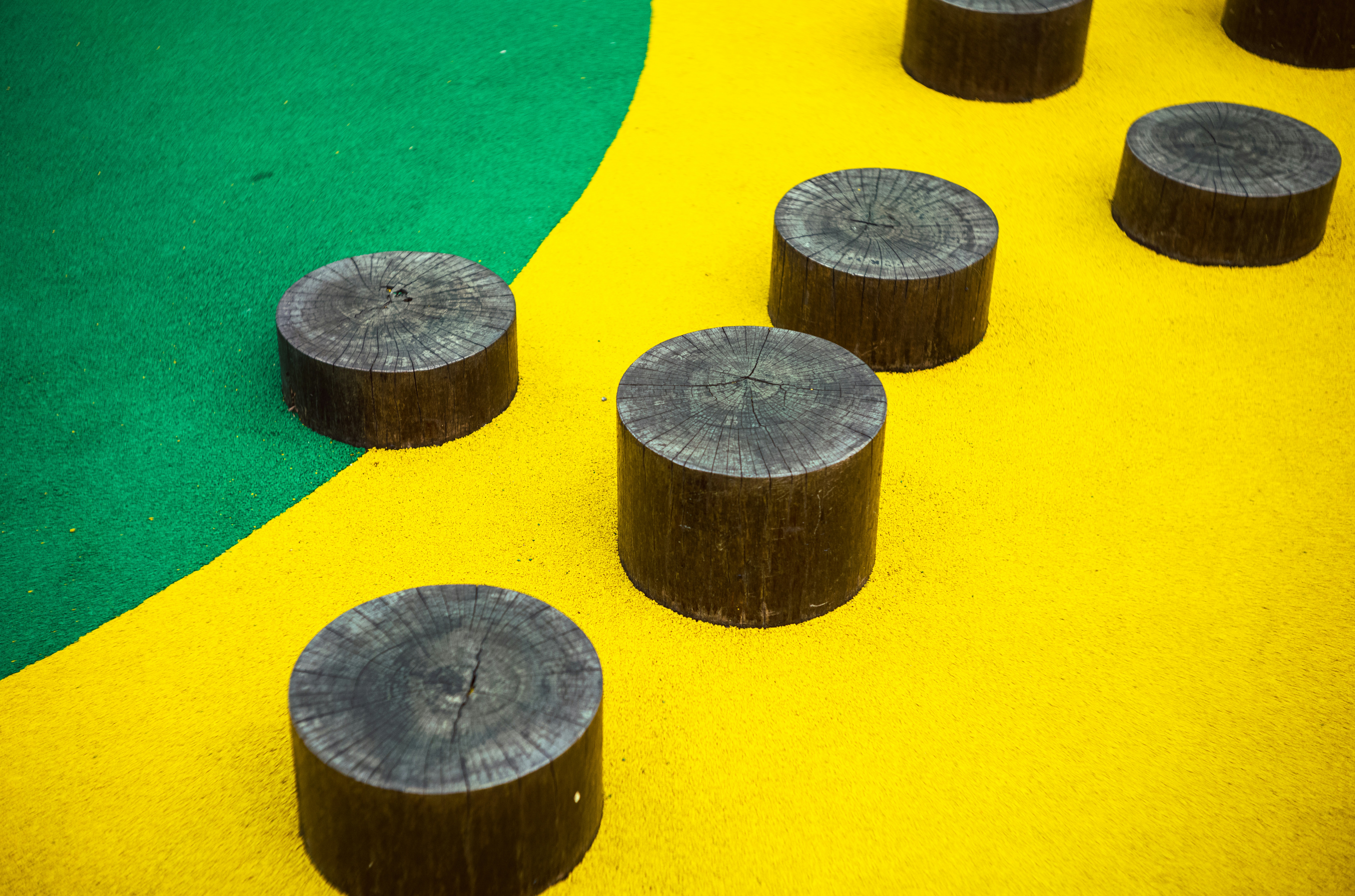 Close-up photograph of dark wooden knobs embedded in a bright yellow playground surface, with a curved green edge in the background. The image emphasizes texture and color contrast.