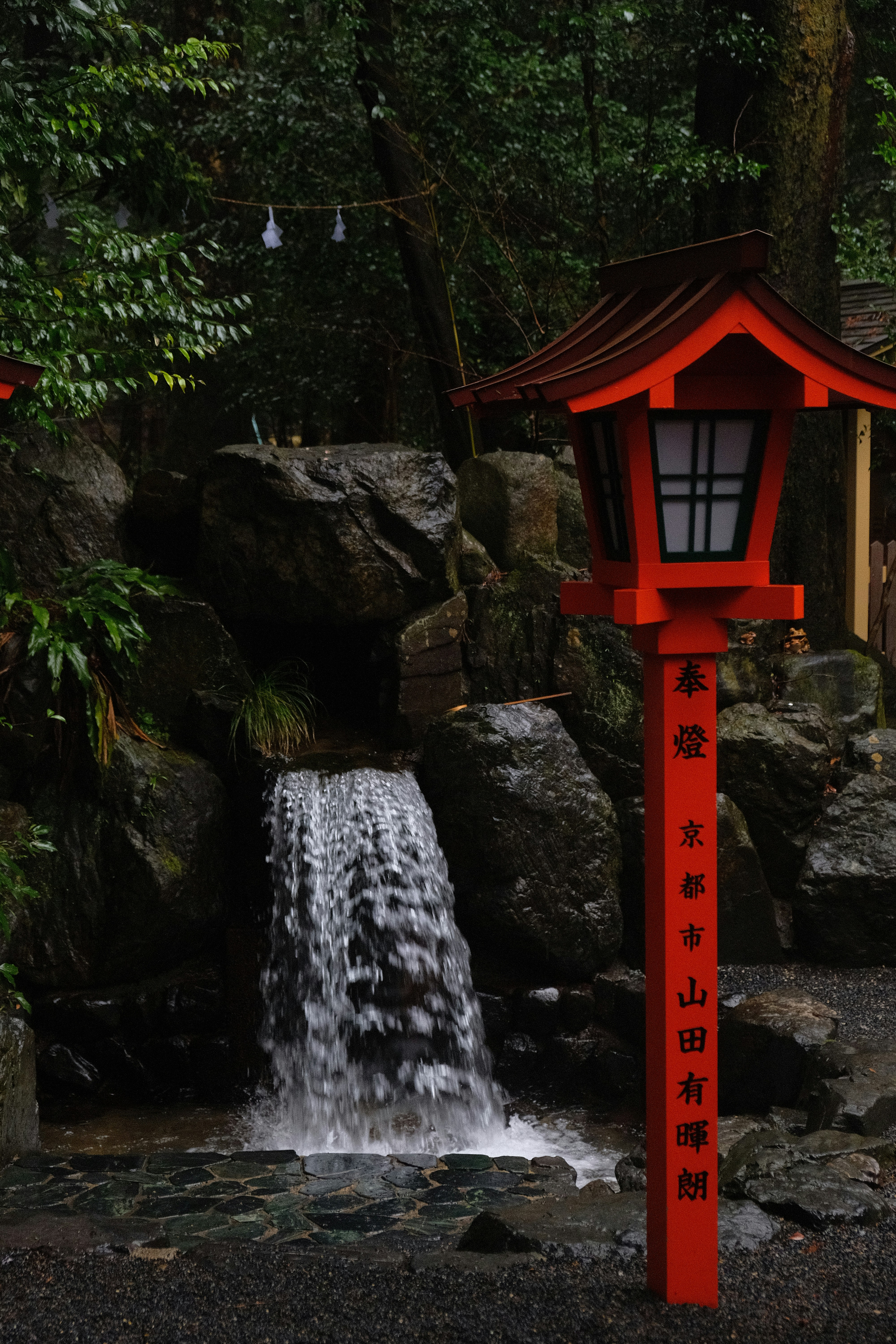 Red lantern stands beside a gentle waterfall in a lush forest setting.
