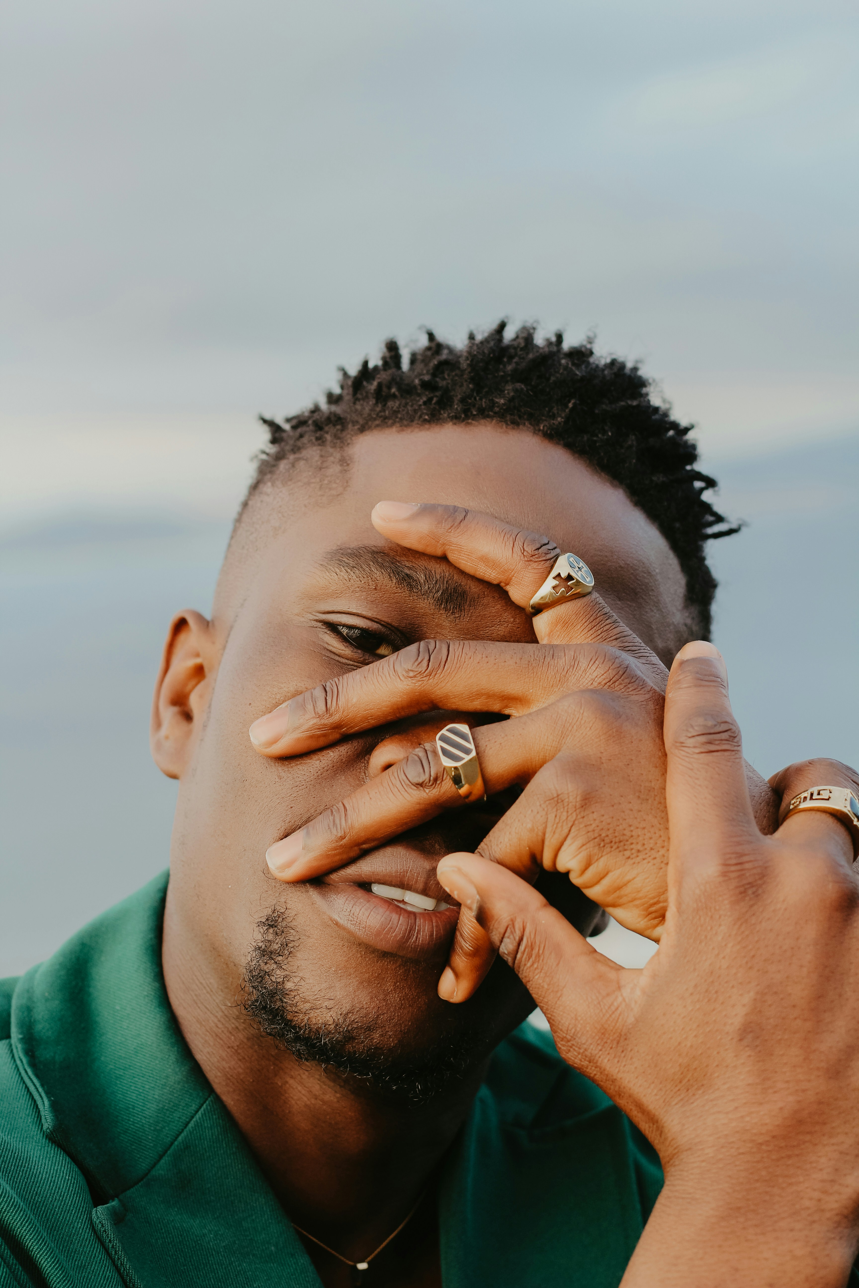 A man partially obscures his face with his hands, adorned with rings, against a softly blurred background. The image evokes a sense of mystery and contemplation.