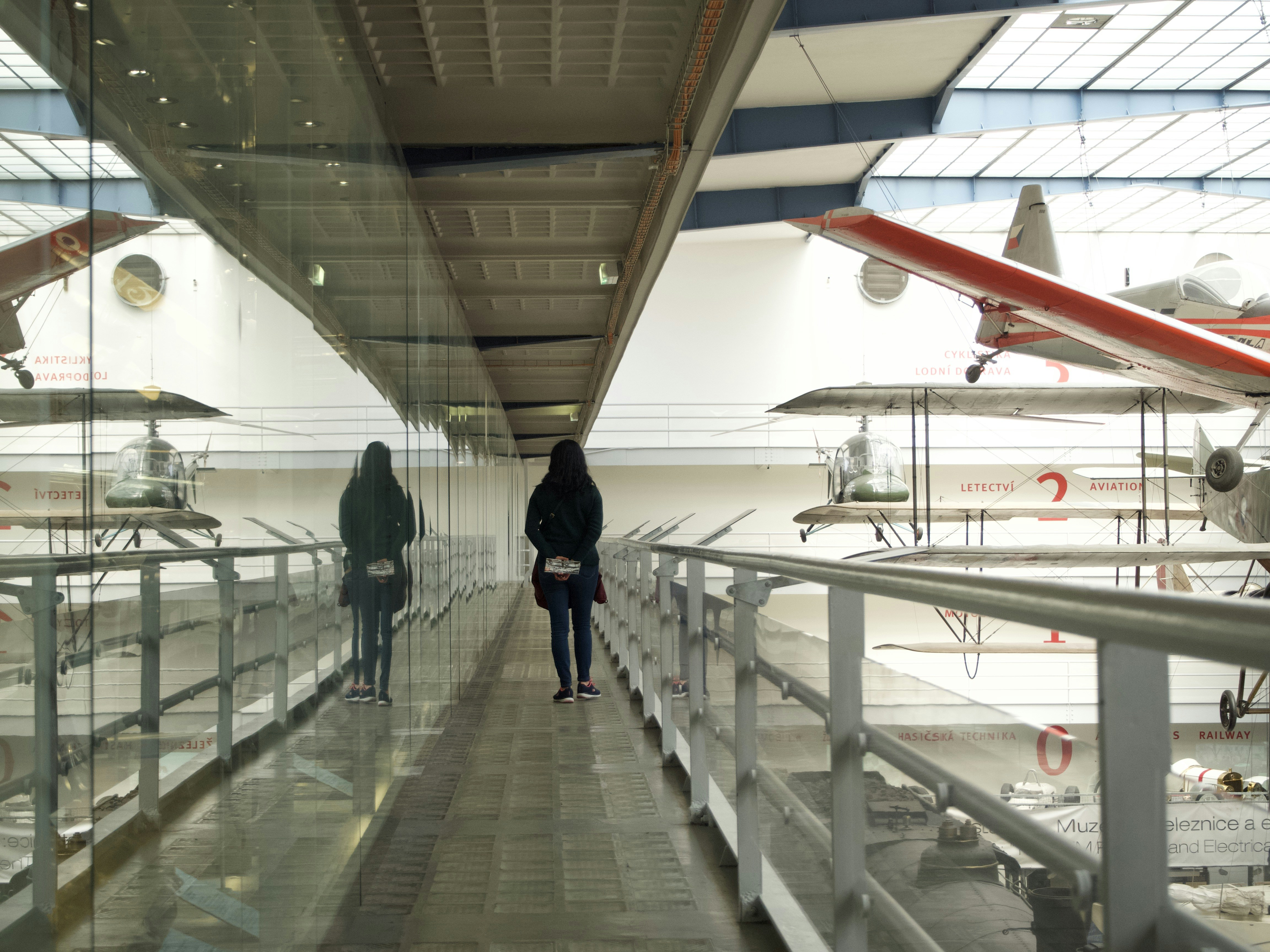 Person walking through an aviation museum with vintage aircraft suspended above, mirrored in the glass wall.