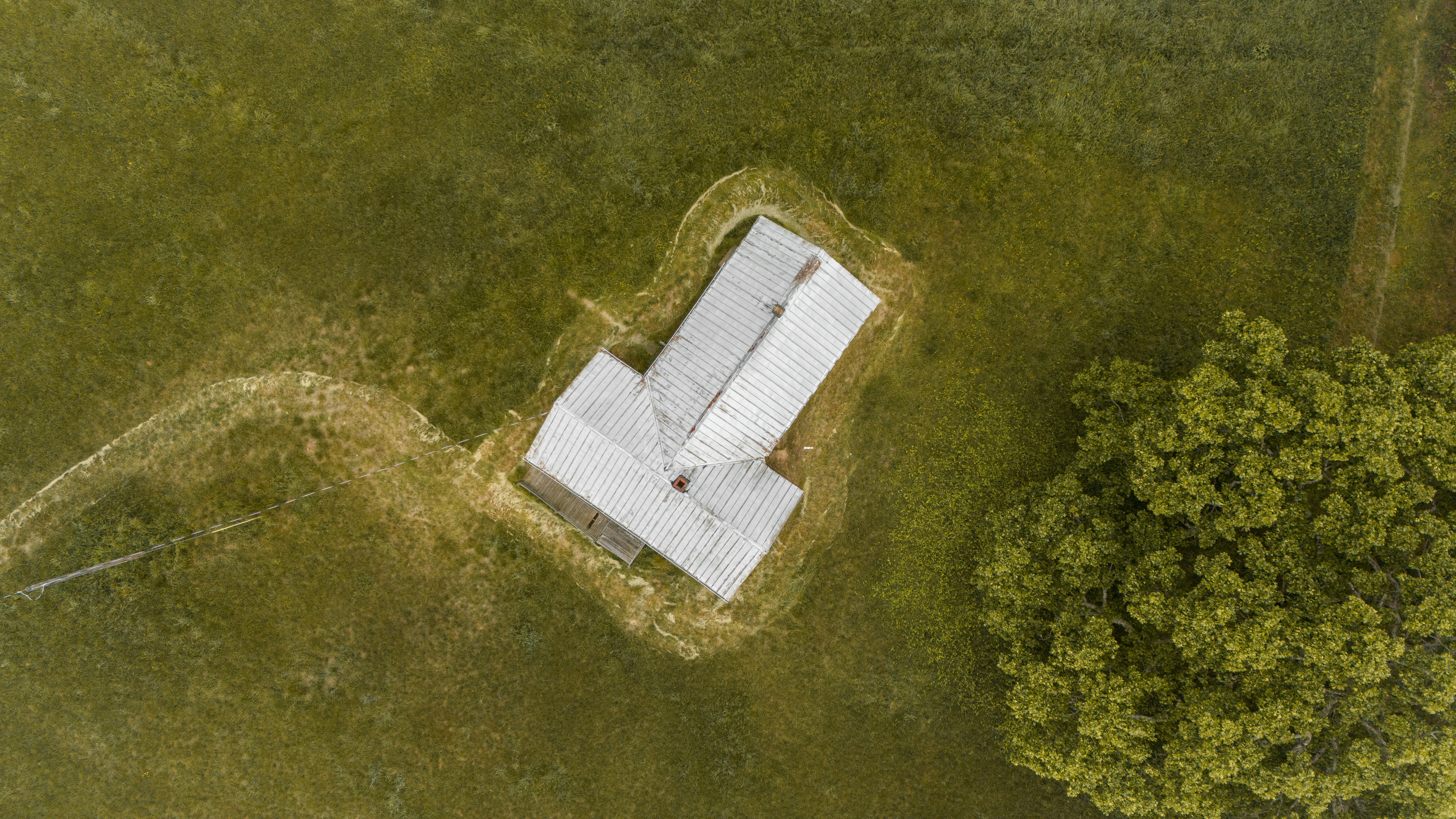 Aerial view of a quaint, isolated homestead surrounded by lush greenery, showcasing a rustic roof and a nearby tree. The landscape conveys a sense of tranquility.