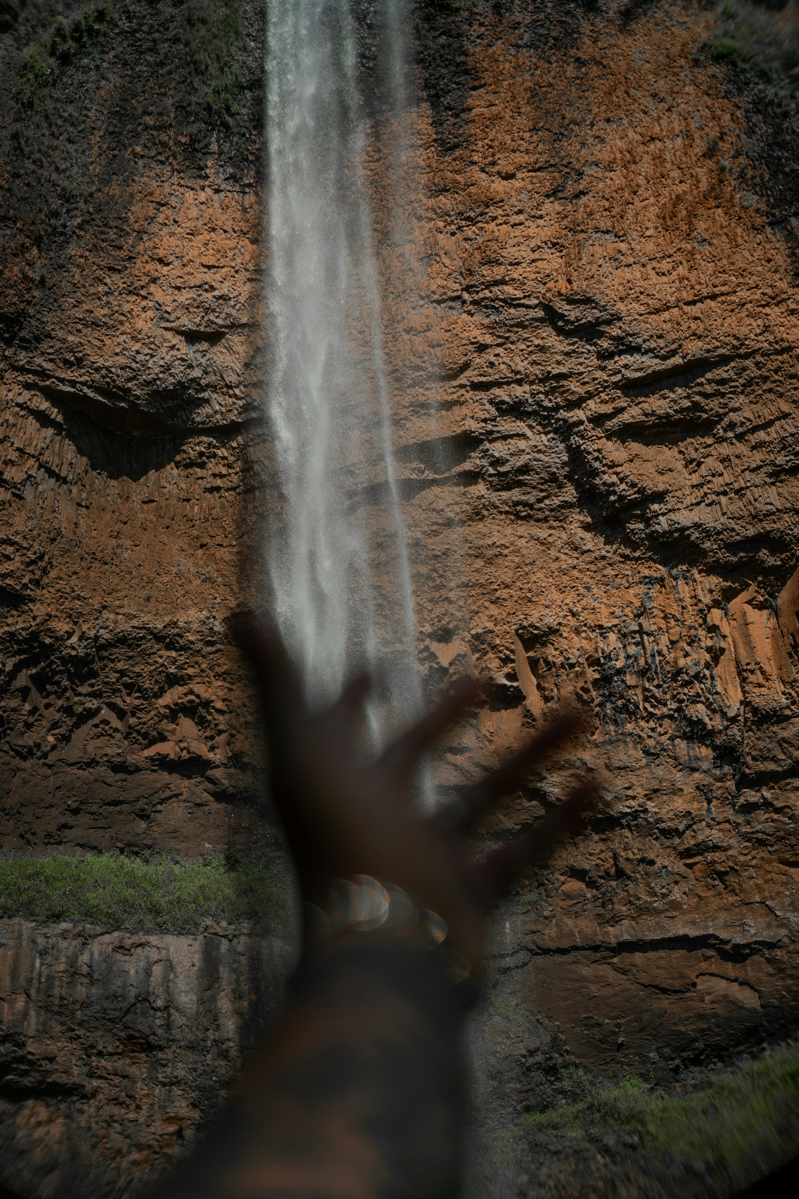 Gesture in the Mist: A Blurred Hand Before a Rust Cliff Waterfall