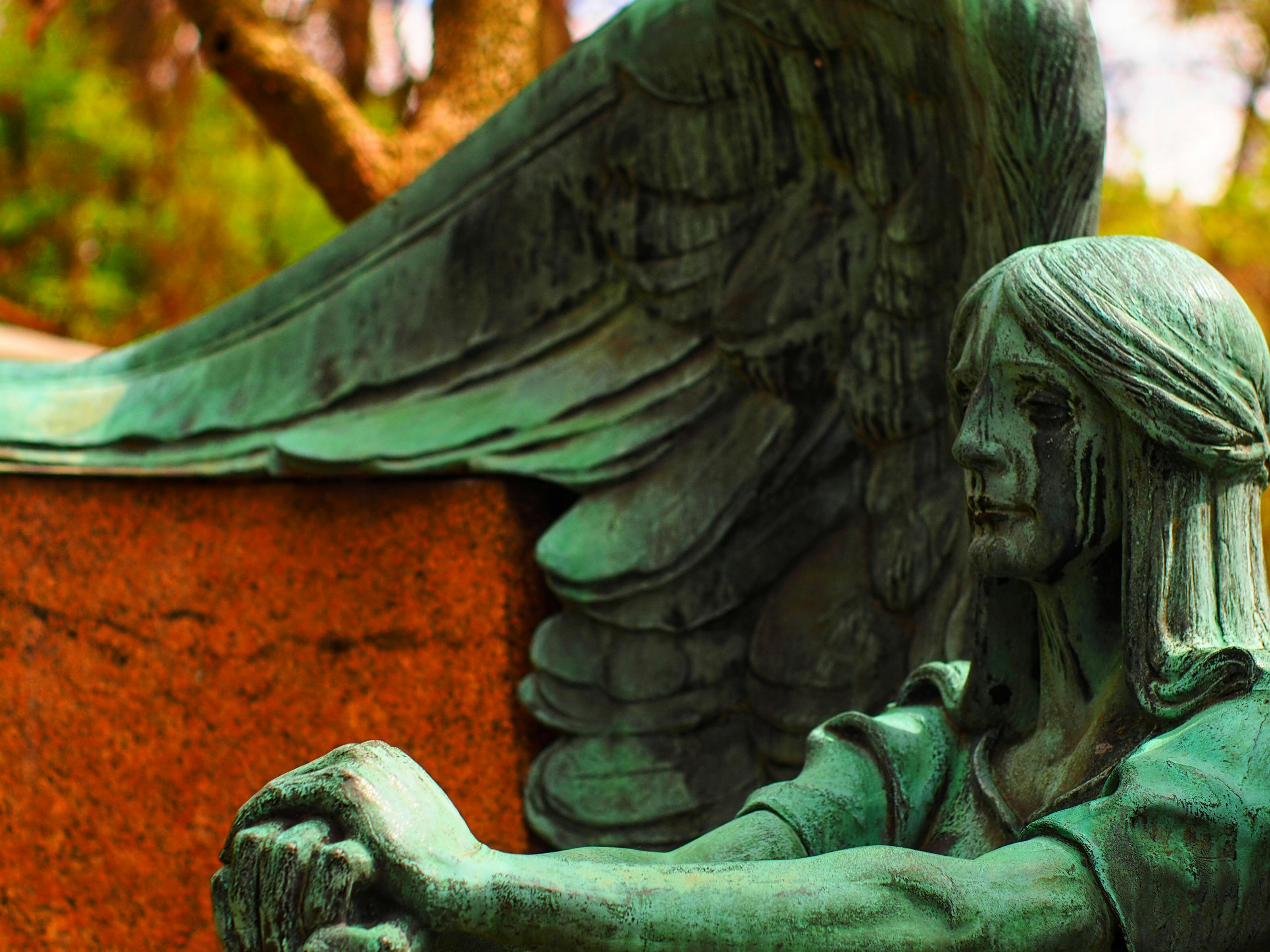 Bronze statue of an angel with folded hands and wings partially visible, set against a blurred natural background.