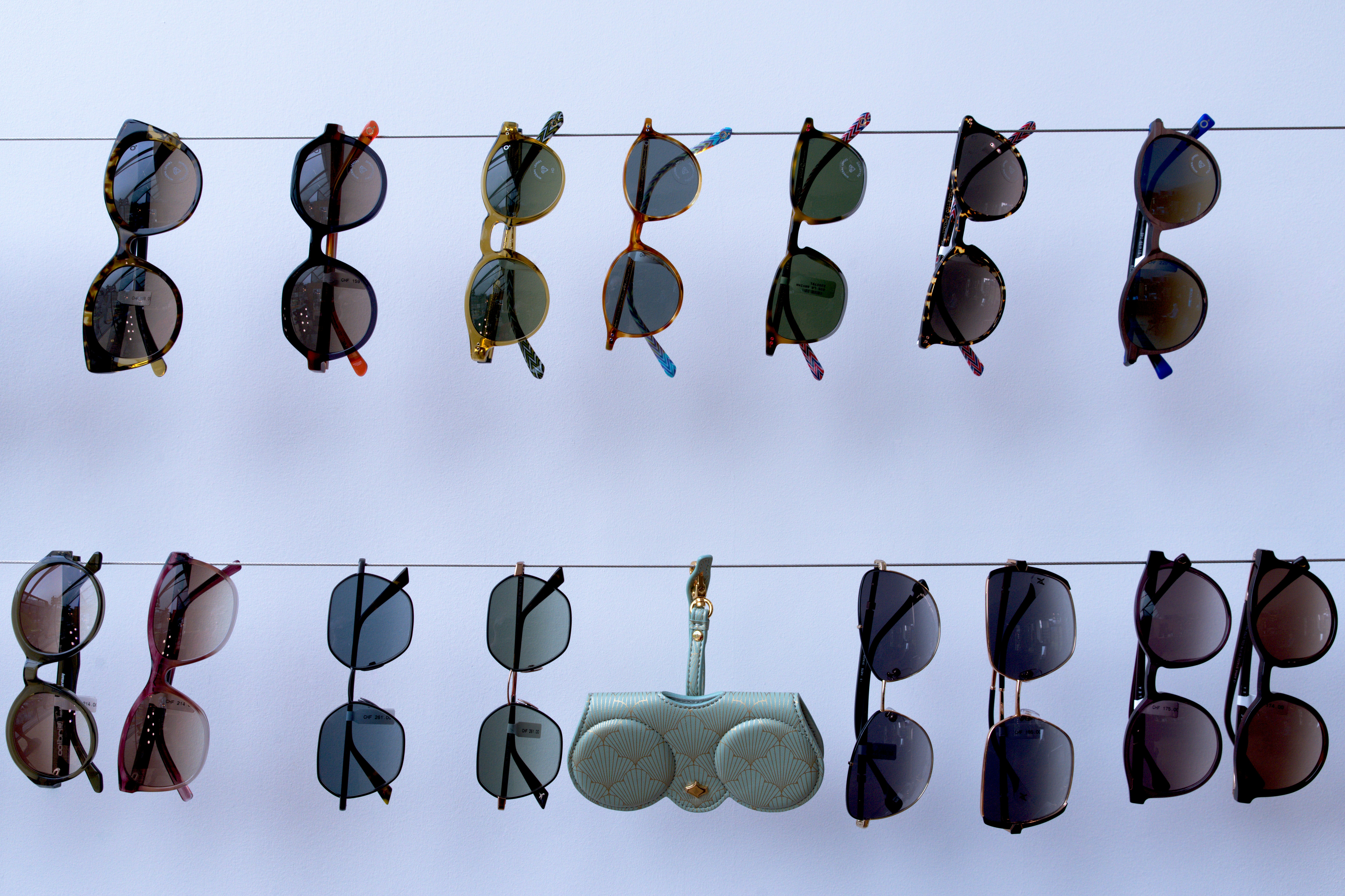 Two parallel rows of sunglasses hang on lines against a pale blue backdrop, with a teal centerpiece centered between them.