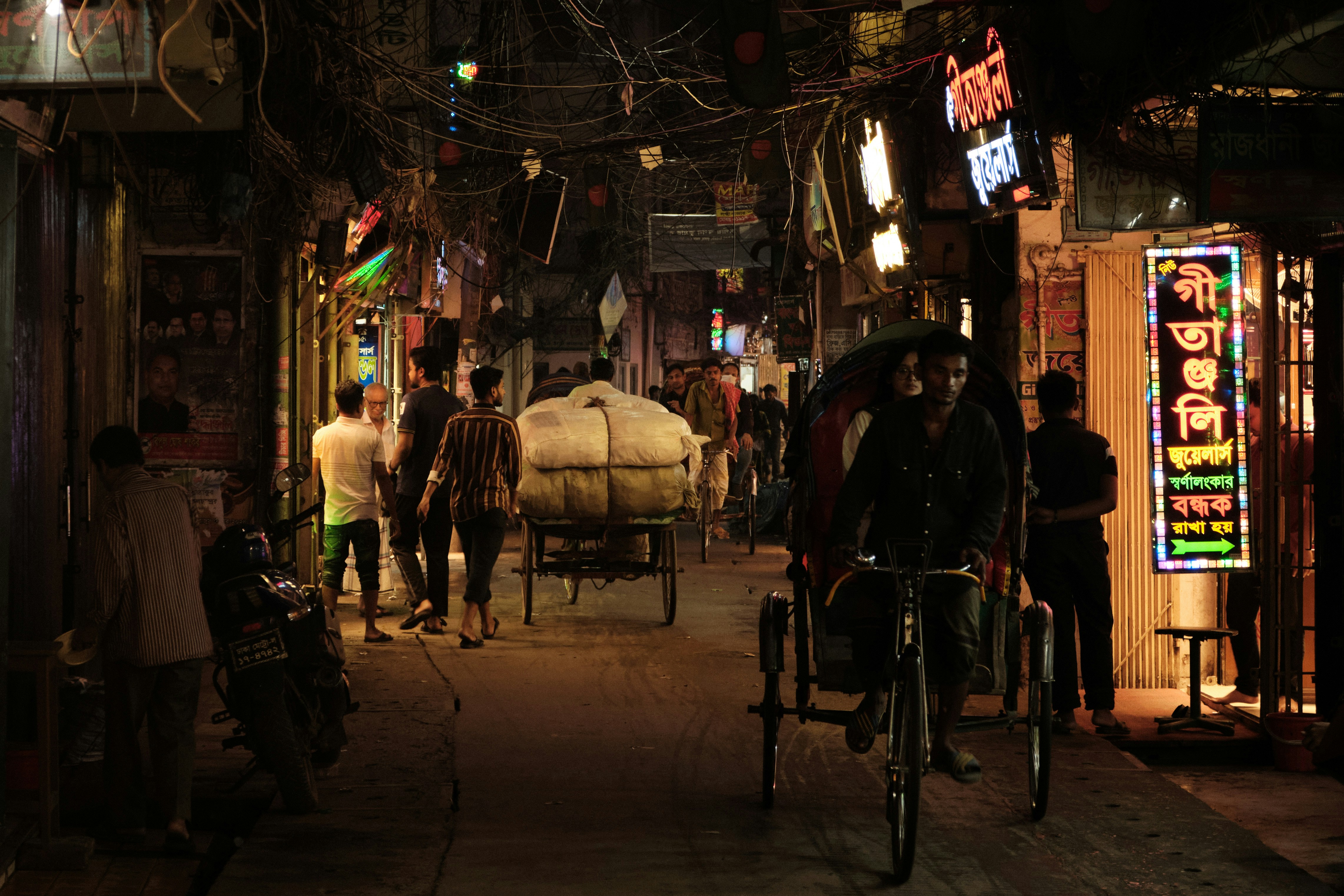 Bustling narrow street in Dhaka with vibrant neon signs and rickshaws navigating the crowd.