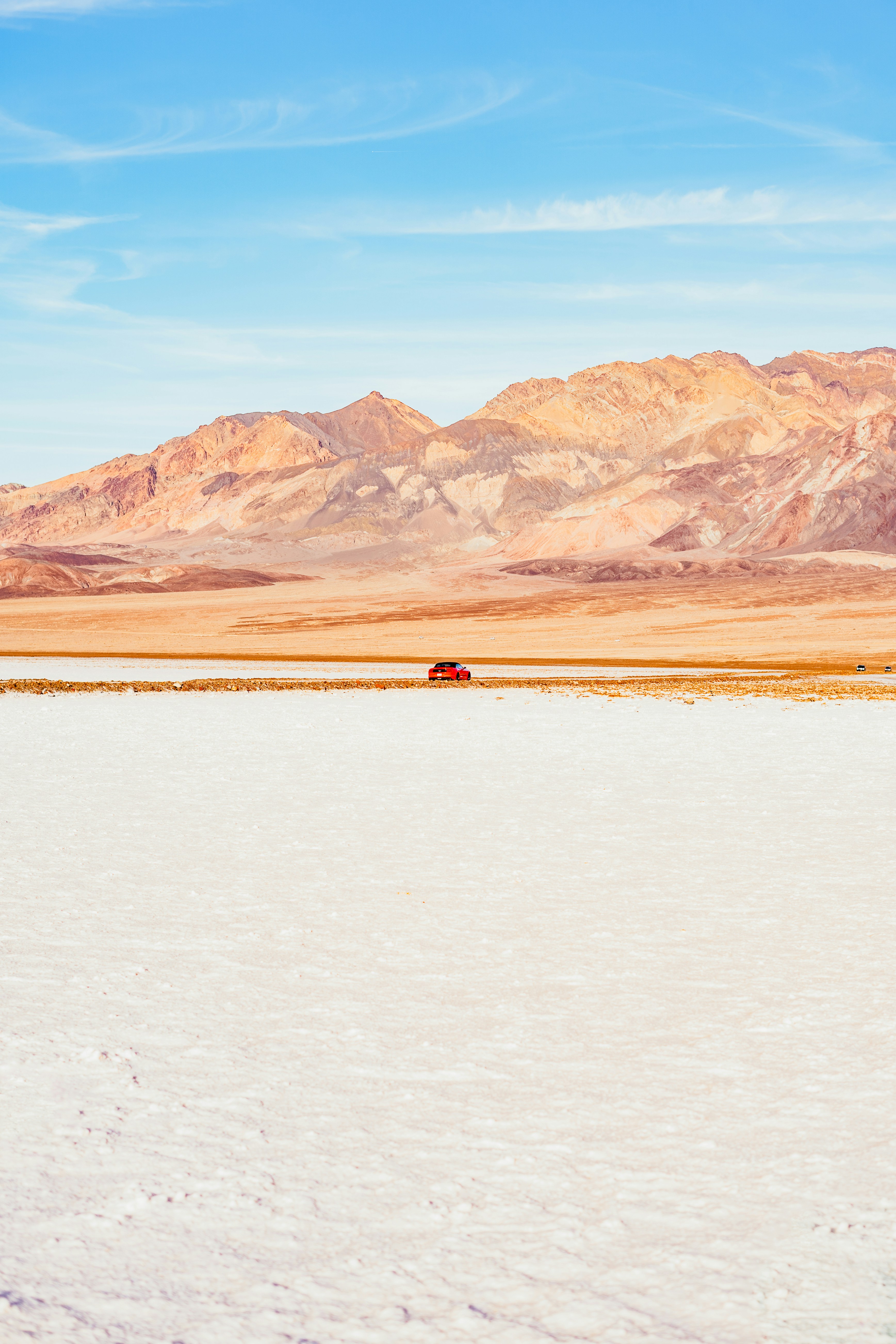 A lone vehicle traversing the vast, white salt flats against a backdrop of rugged mountains under a clear blue sky.