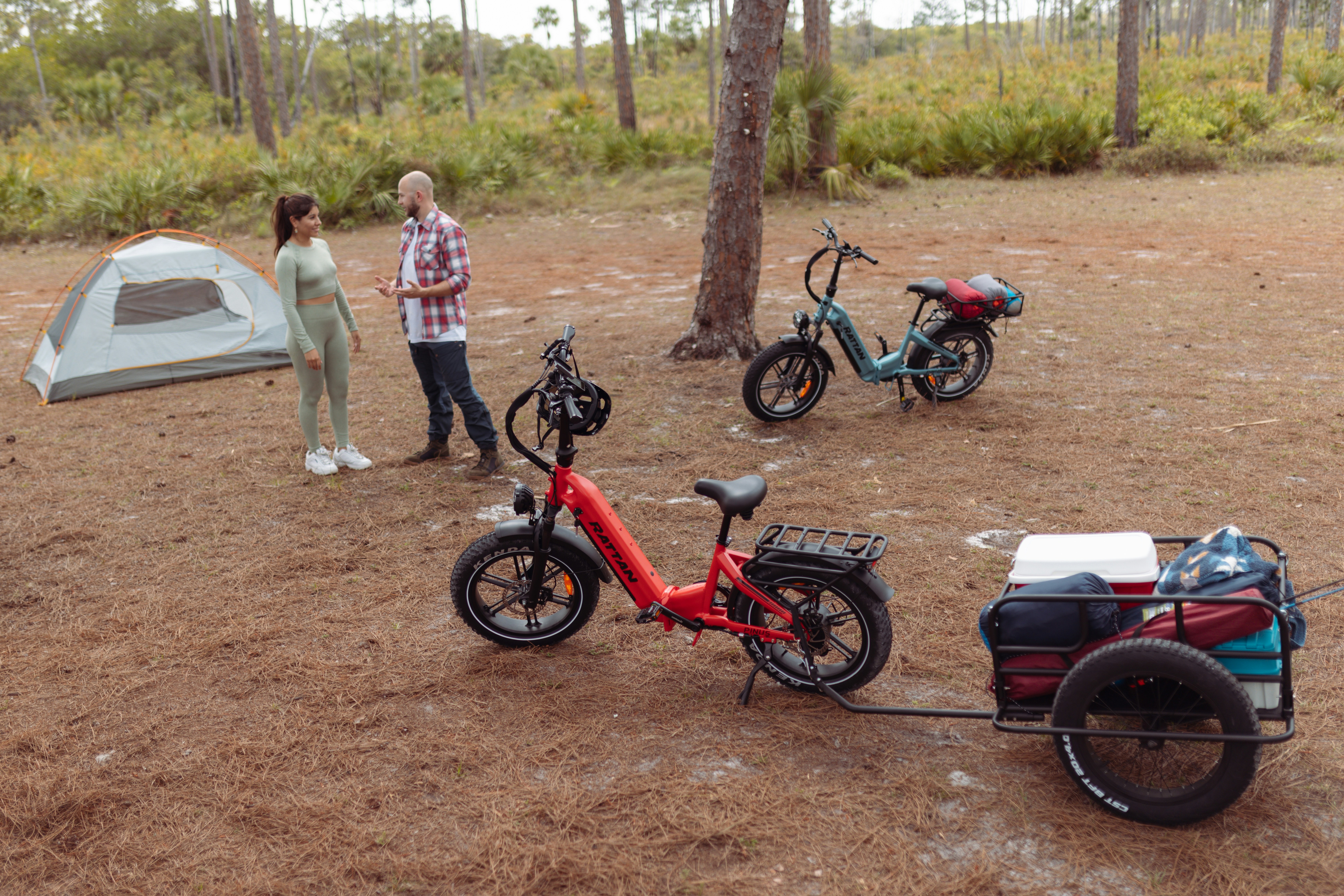 Couple in a campsite with red electric scooter and trailer under pines