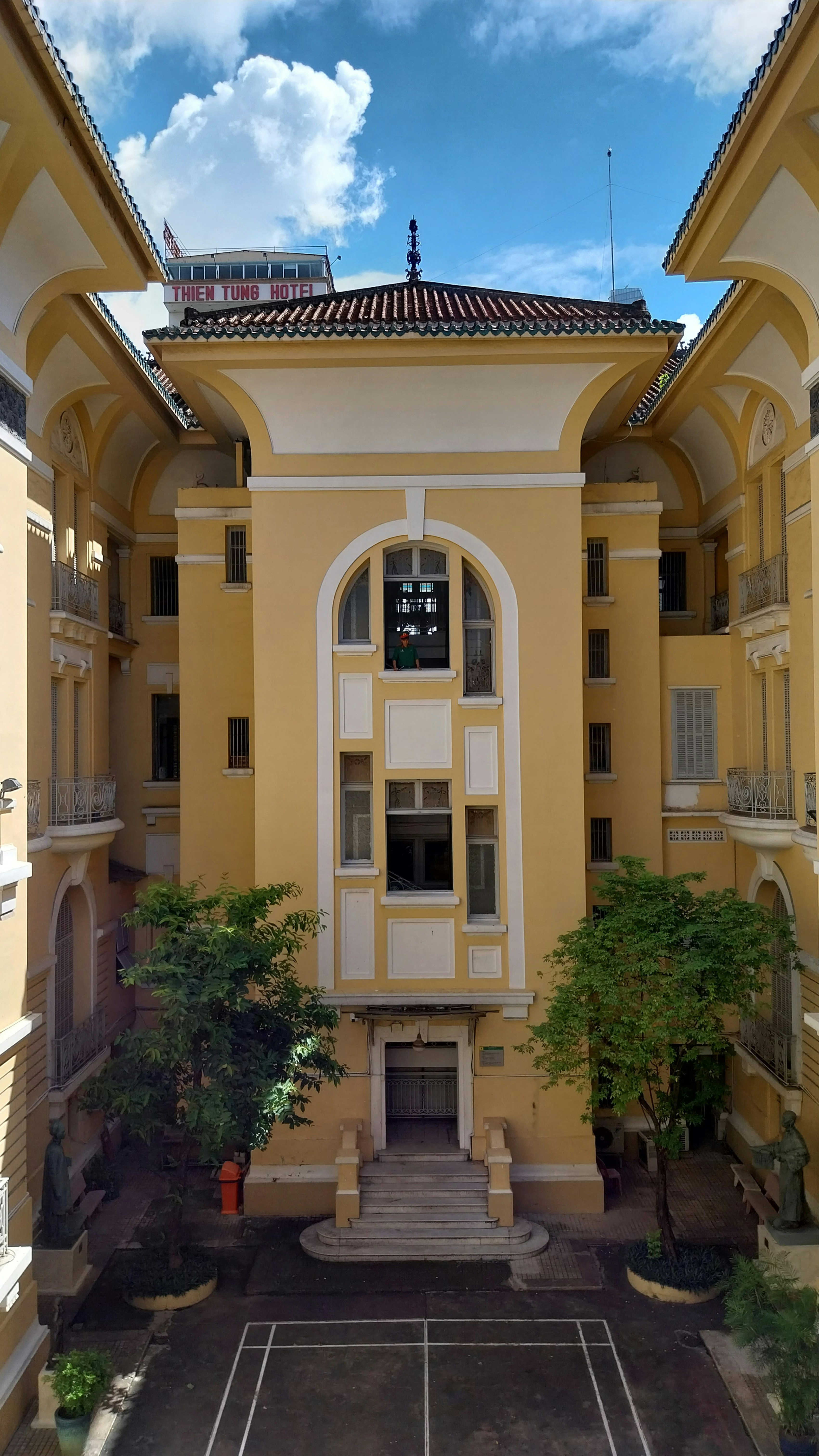 Photograph of a sunlit yellow courtyard building with a central tower and arched windows, framed by symmetrical wings and greenery.