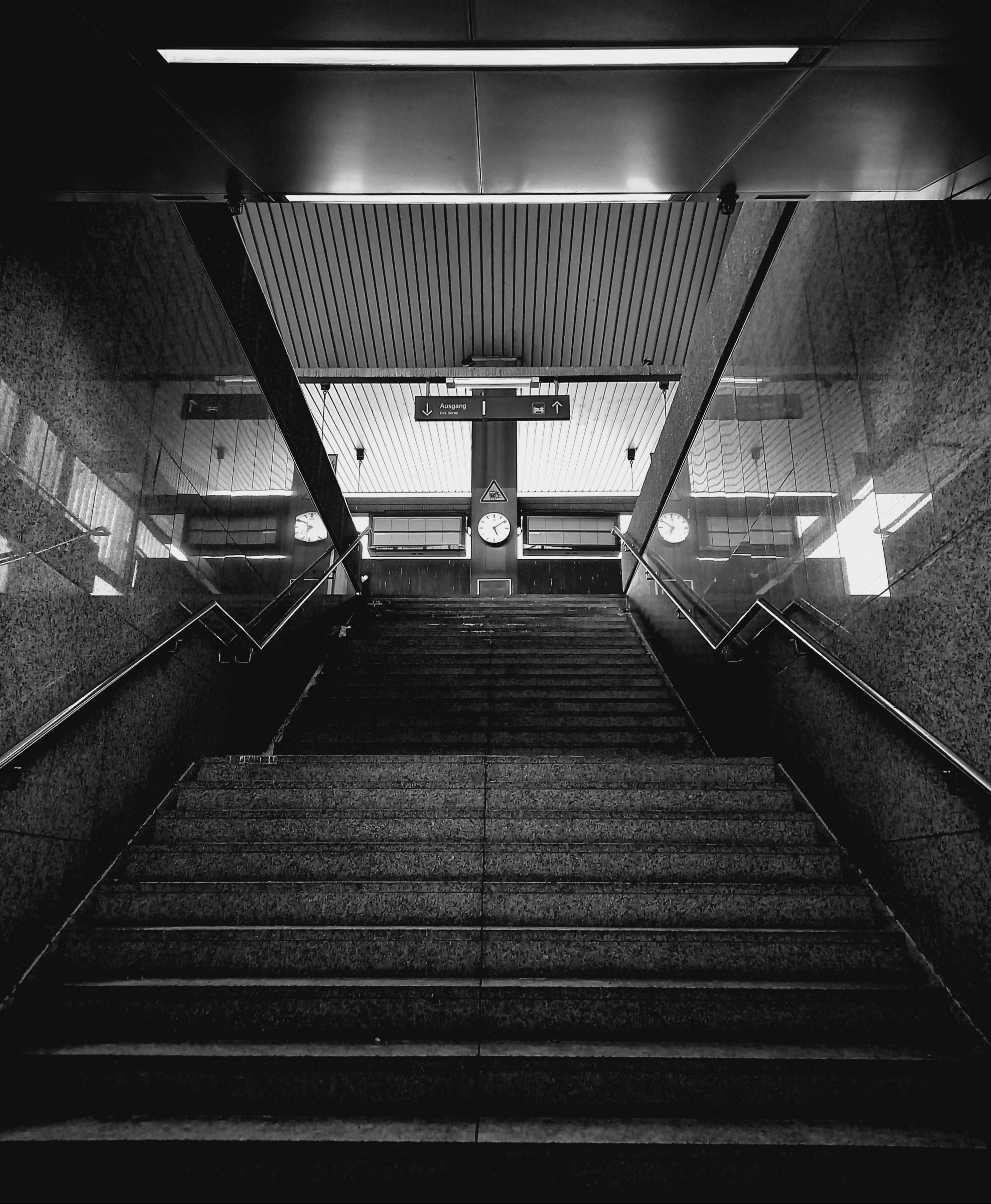 Symmetrical subway stairs ascend to a central clock, framed by glossy walls and overhead signage in stark monochrome.