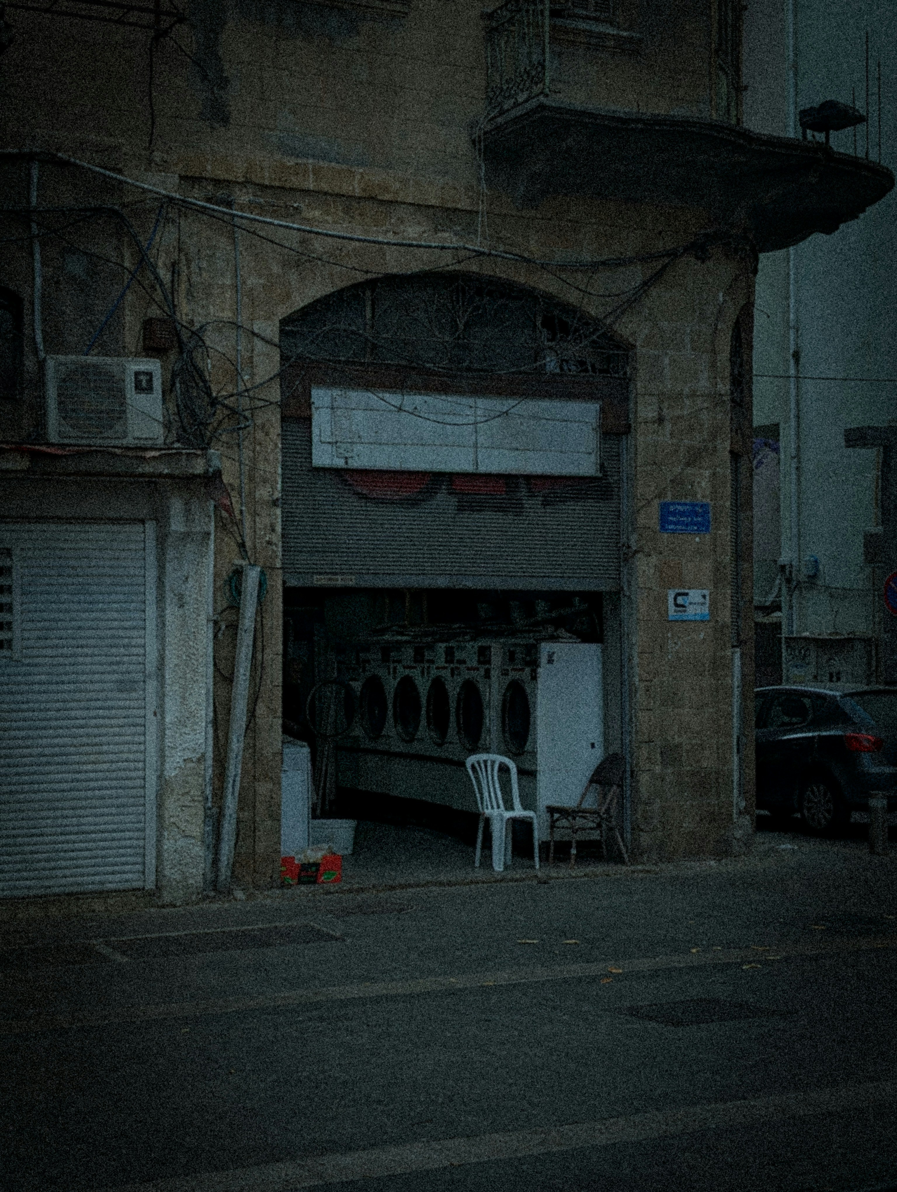 Dim street at night reveals an open laundromat with visible washing machines and a lone white chair outside. The moody, blue-lit storefront captures a quiet urban moment.