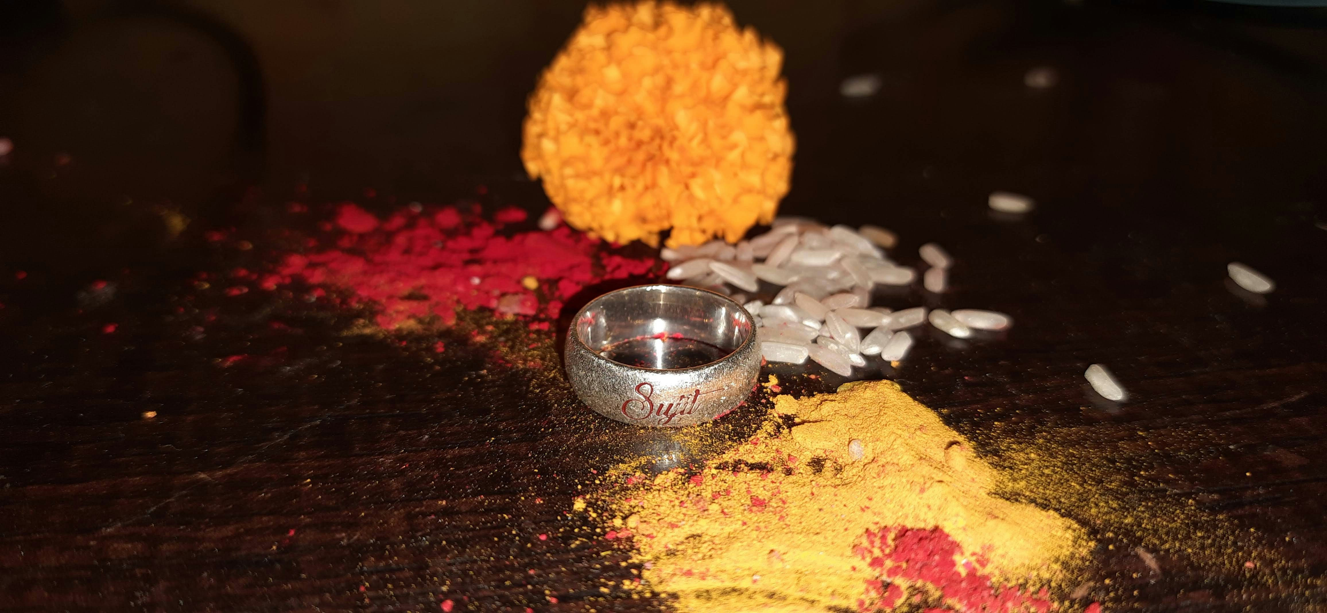 A silver ring sits on a dark wooden surface, surrounded by red and yellow pigment spills and scattered white stones. An orange bloom adds a warm contrast to the scene.