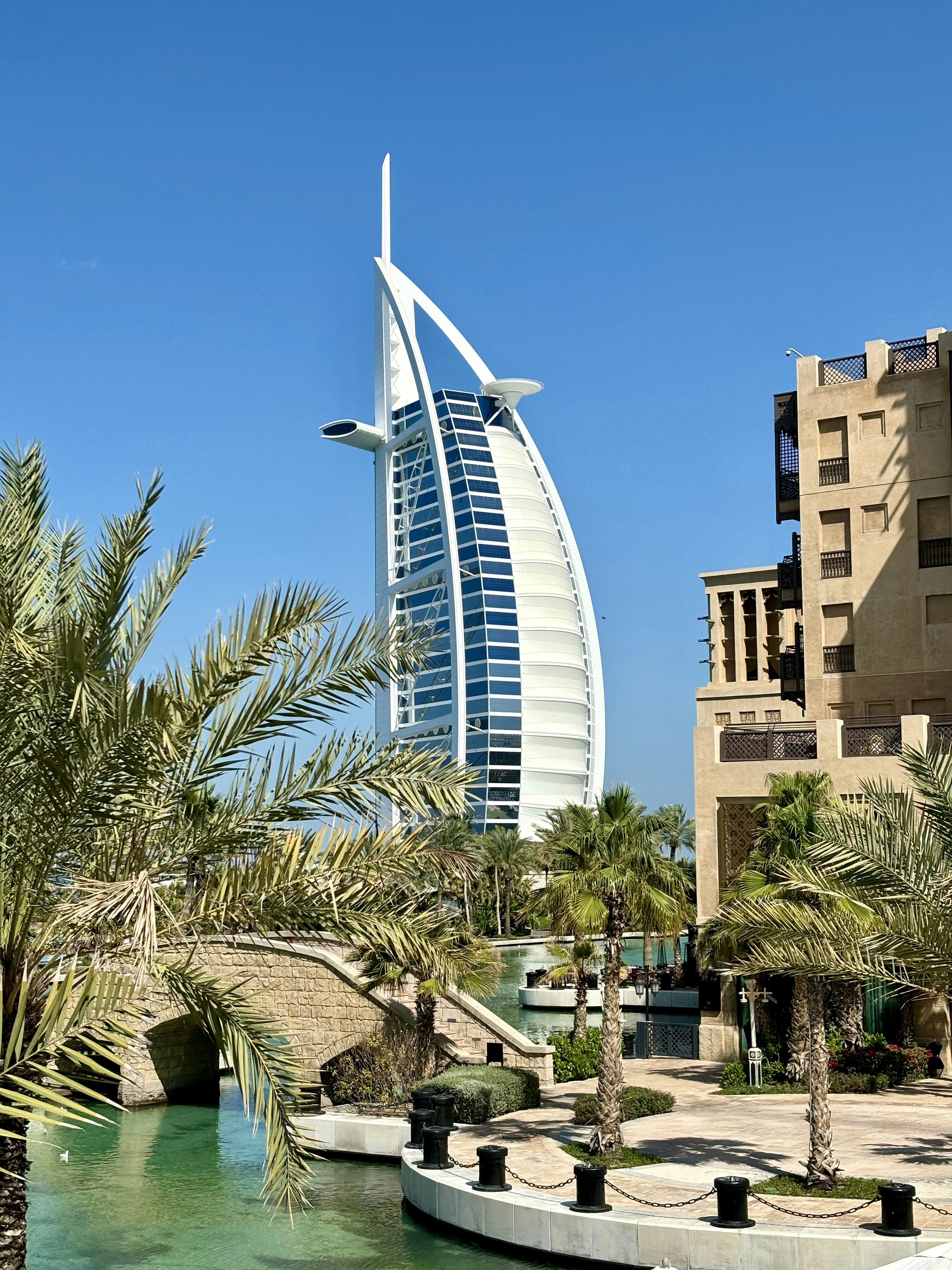 Iconic Burj Al Arab rising majestically beside traditional Middle Eastern structures, framed by lush palm trees and tranquil waters.
