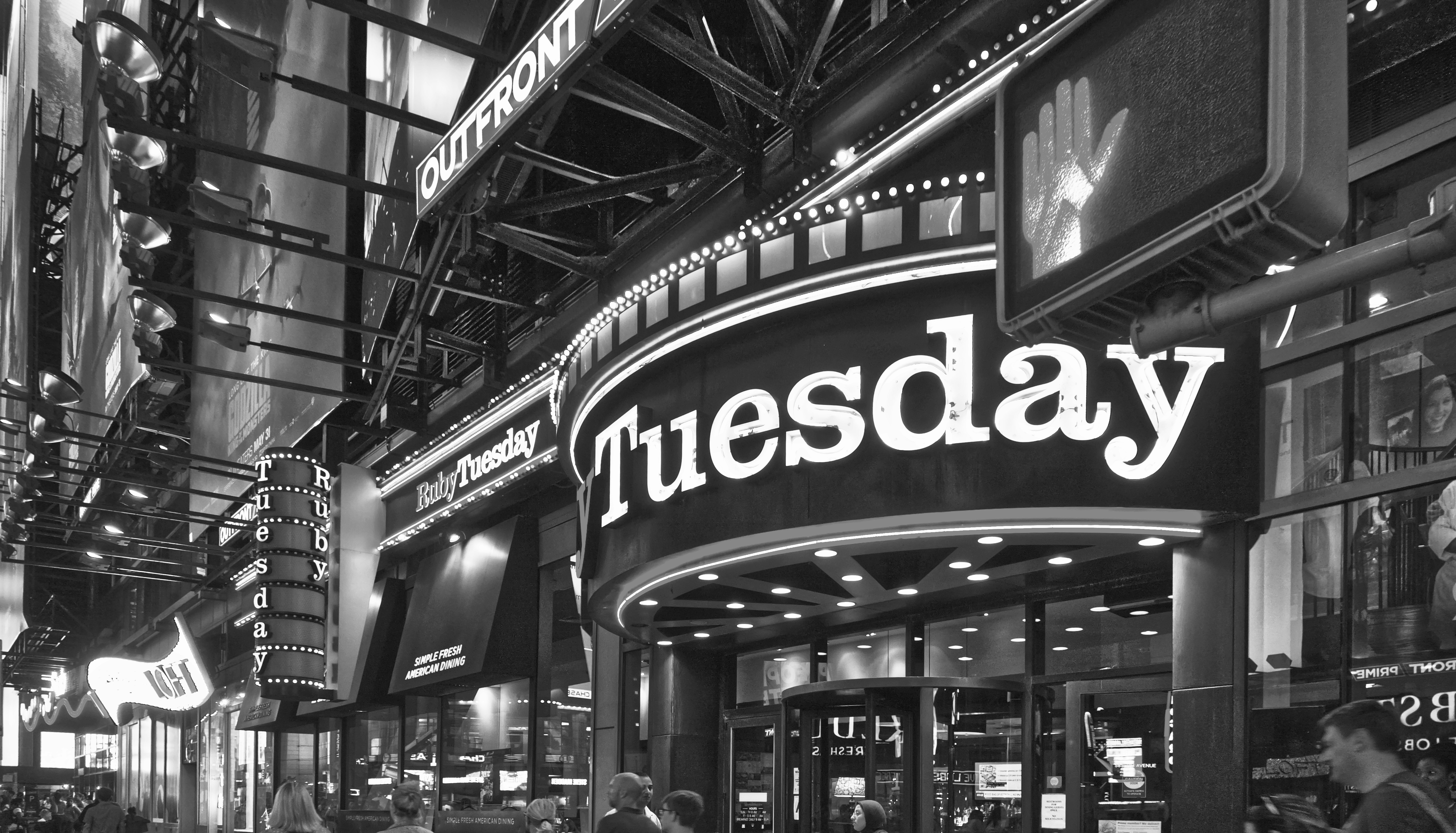 Grayscale street photography photograph capturing a prominent neon marquee reading Tuesday amid a busy urban night scene.