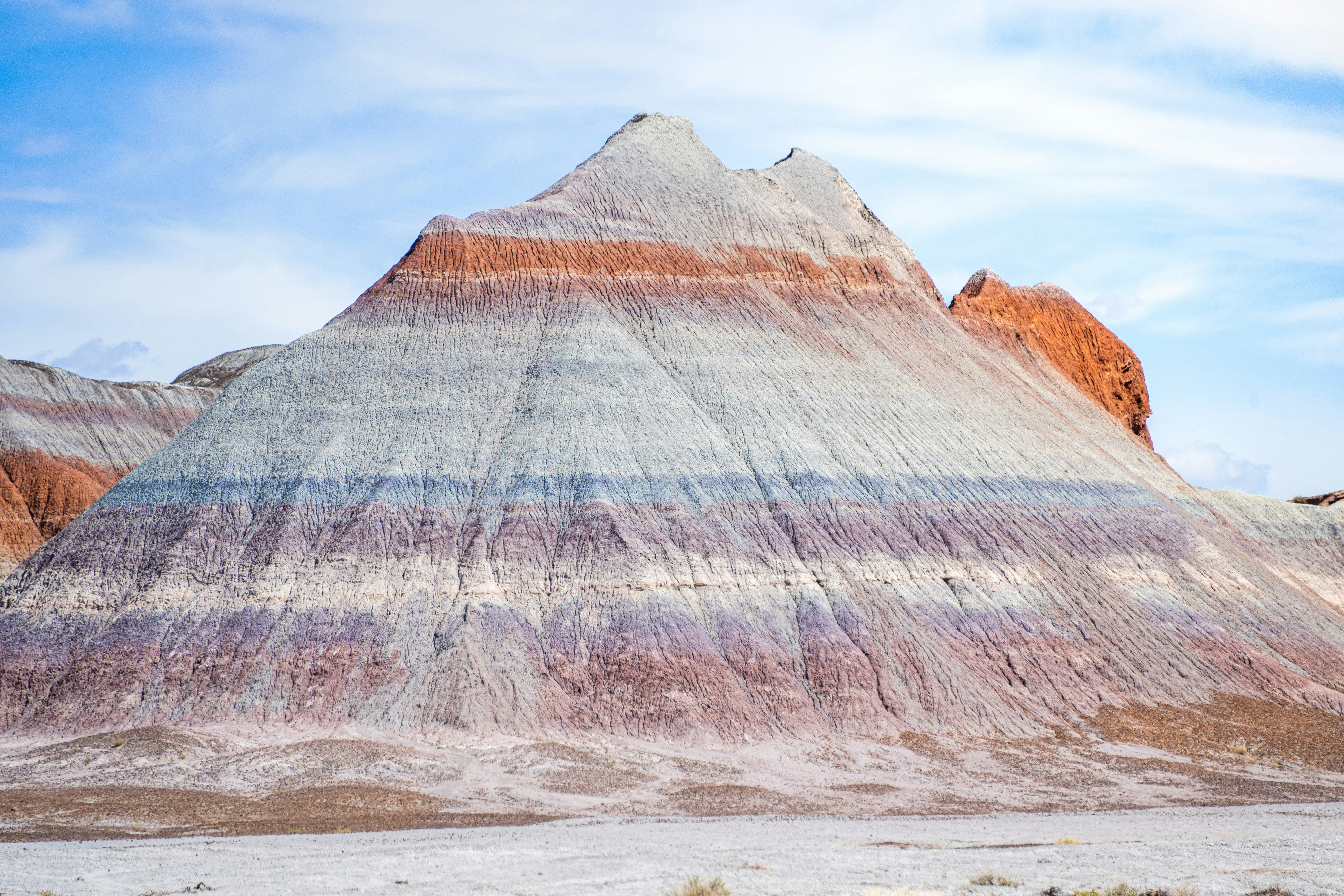 Layered badlands rise from a flat desert, displaying pastel bands of white, gray, blue, purple, and orange under a bright sky. The scene emphasizes sculpted strata and vast openness.